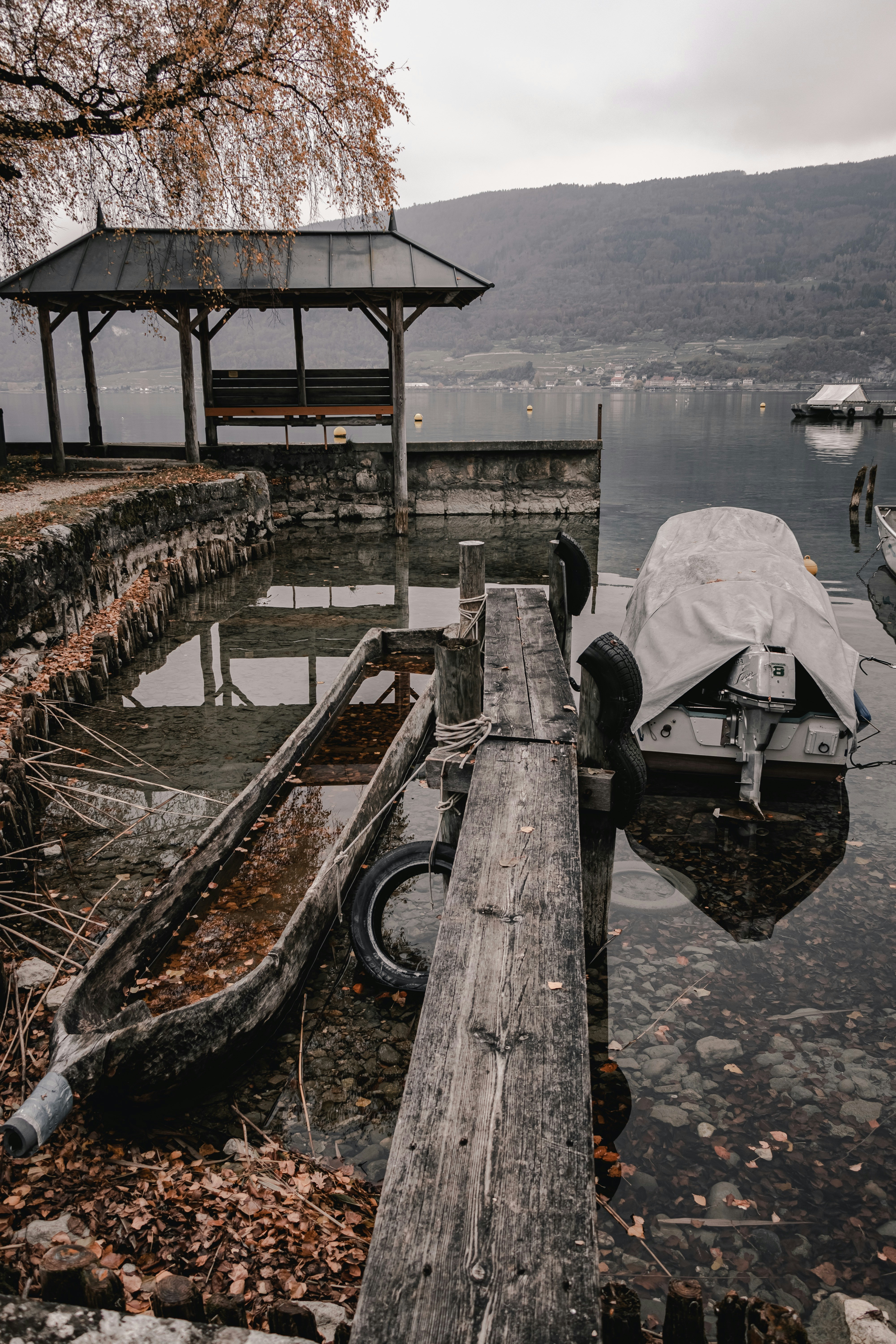 Abandoned wooden boat resting by a tranquil dock, with a covered boat and autumn foliage reflected in the still water.