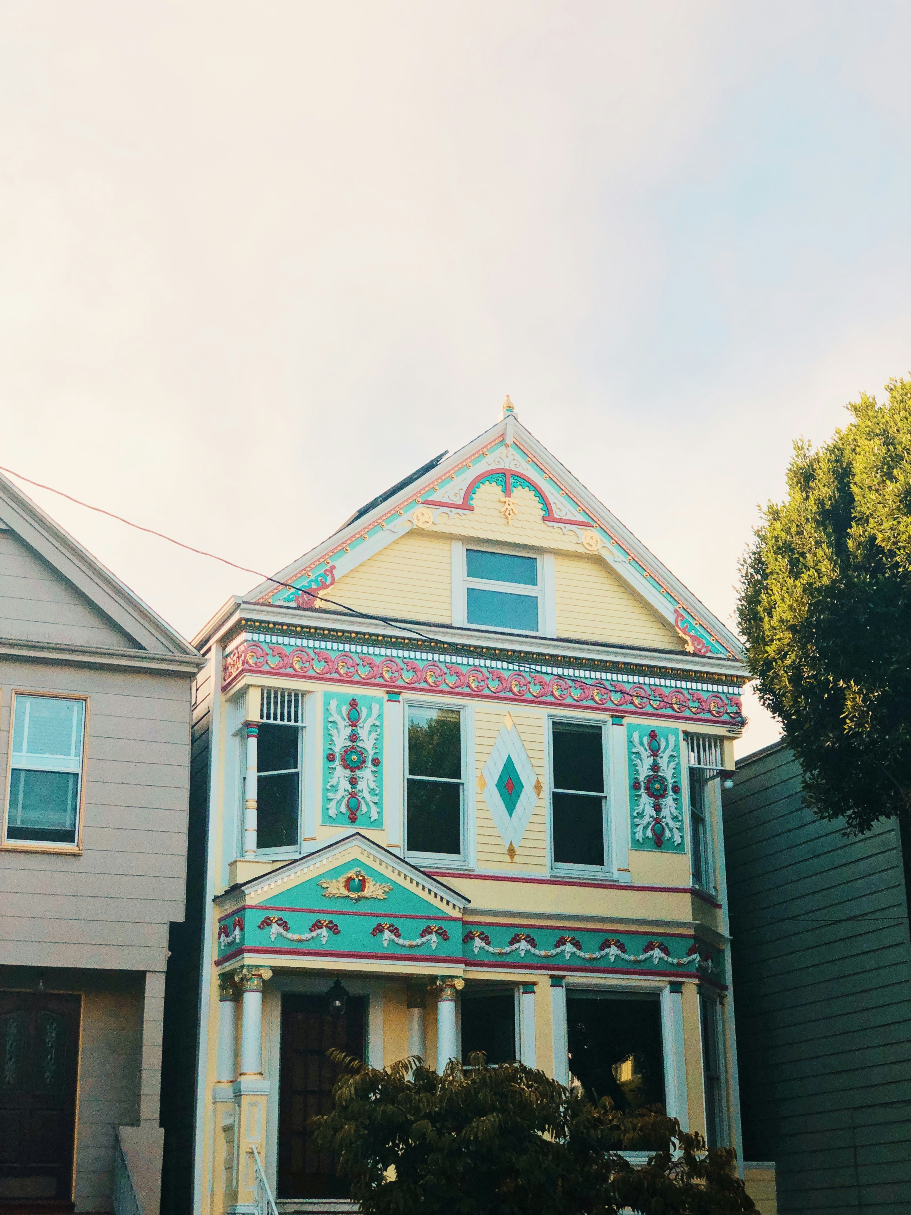 Colorful Victorian house adorned with intricate patterns and designs, nestled between two simpler structures. A lush shrub adds to the charm.