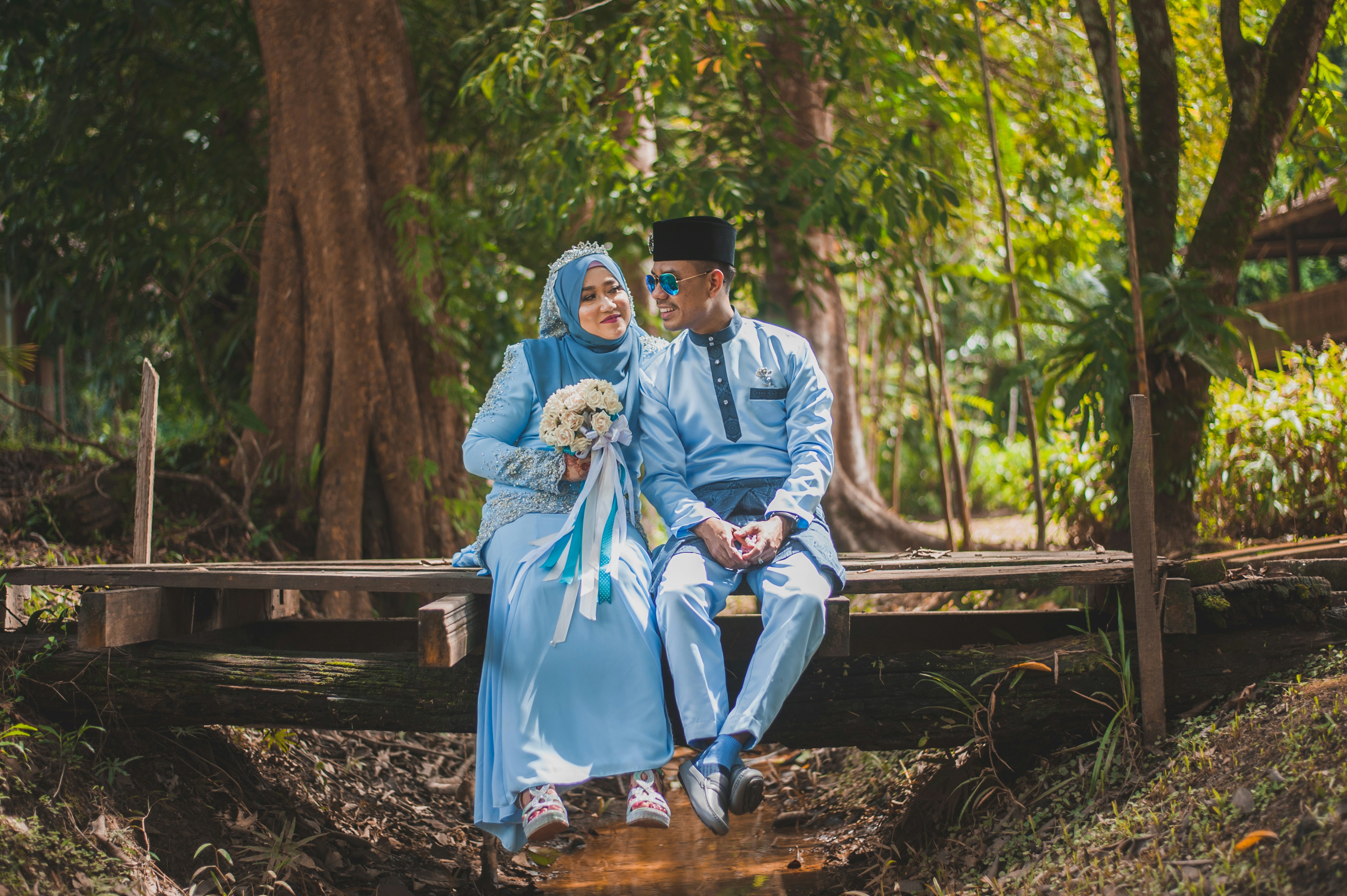 Couple in matching blue attire sitting on a rustic wooden bridge surrounded by lush greenery.
