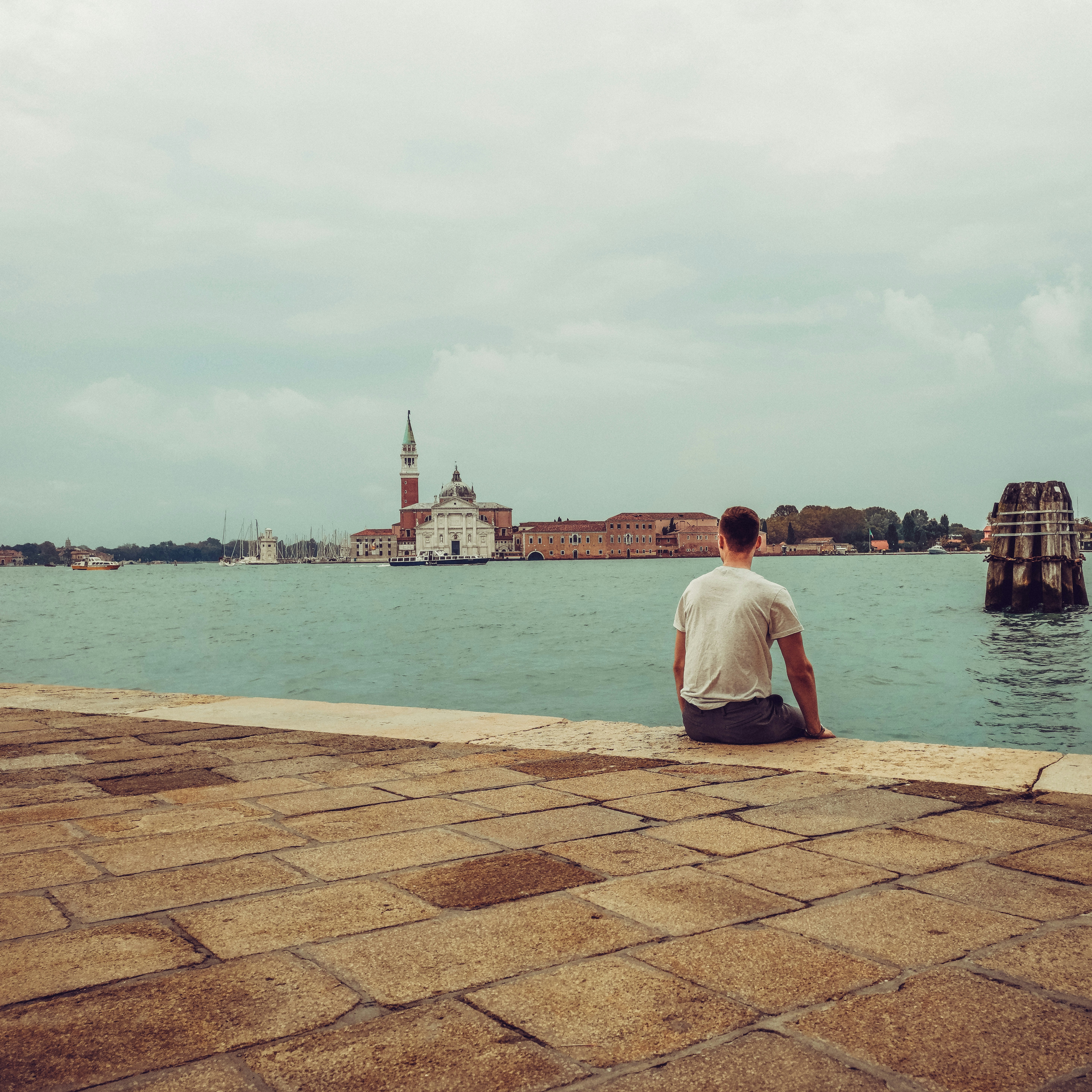 A solitary man sits on a stone quay facing the calm water, with Venice's historic skyline rising across the lagoon. The composition emphasizes solitude and the quiet intersection of land and water.