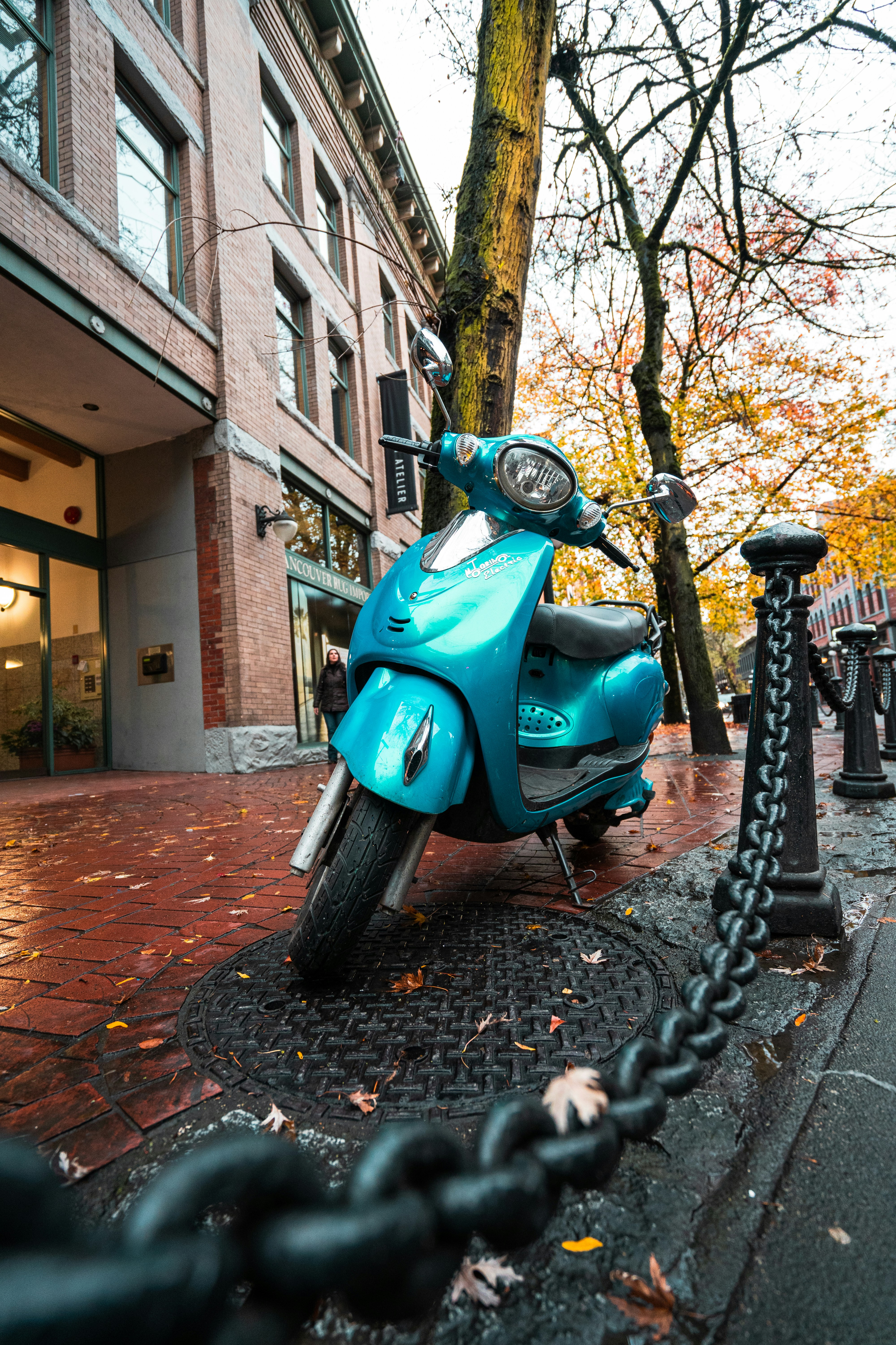 Teal scooter parked on a rainy street, surrounded by autumn leaves and historic brick buildings. A chain fence adds to the urban ambiance.