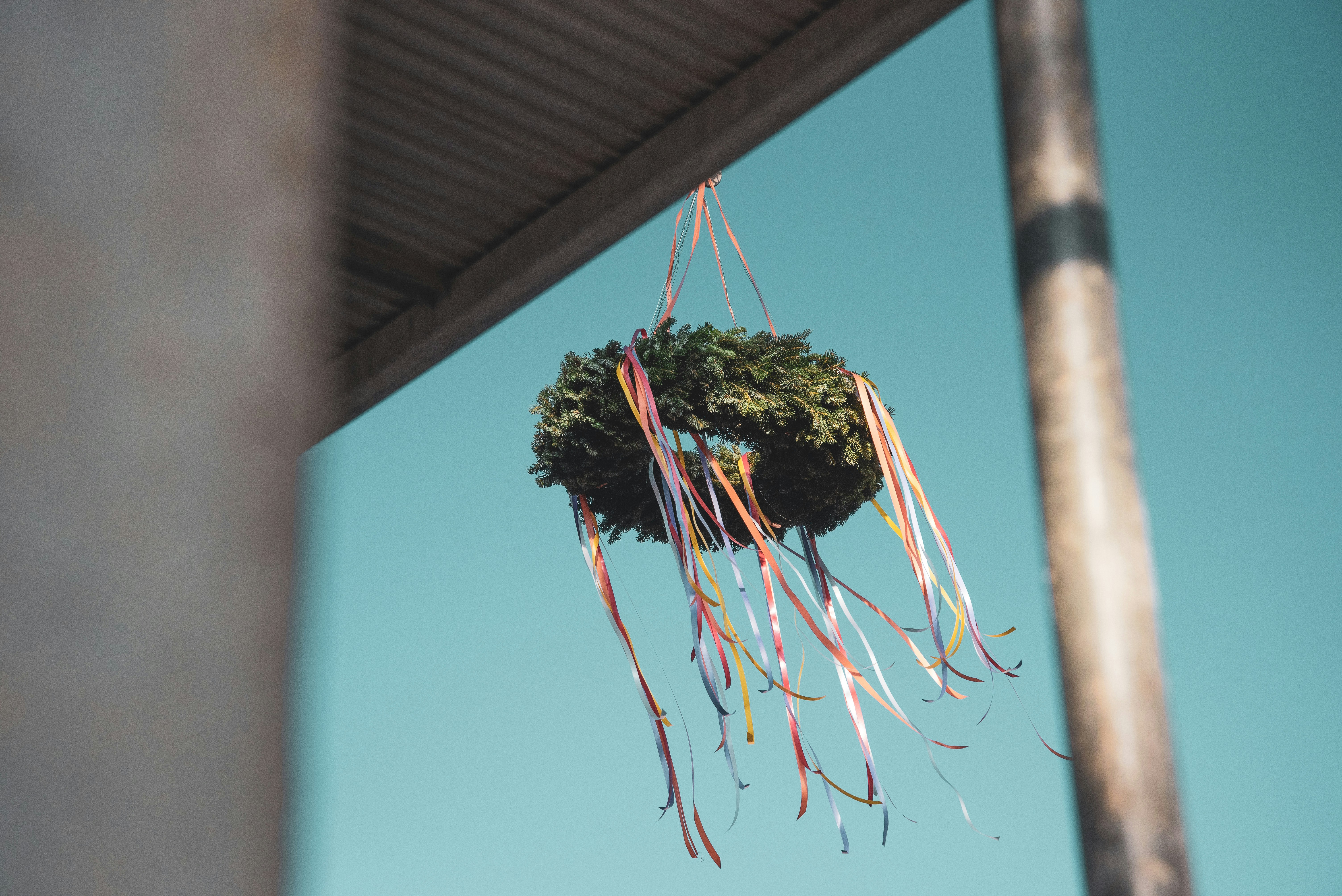 Circular green wreath adorned with flowing ribbons, suspended under a metal roof against a clear sky.