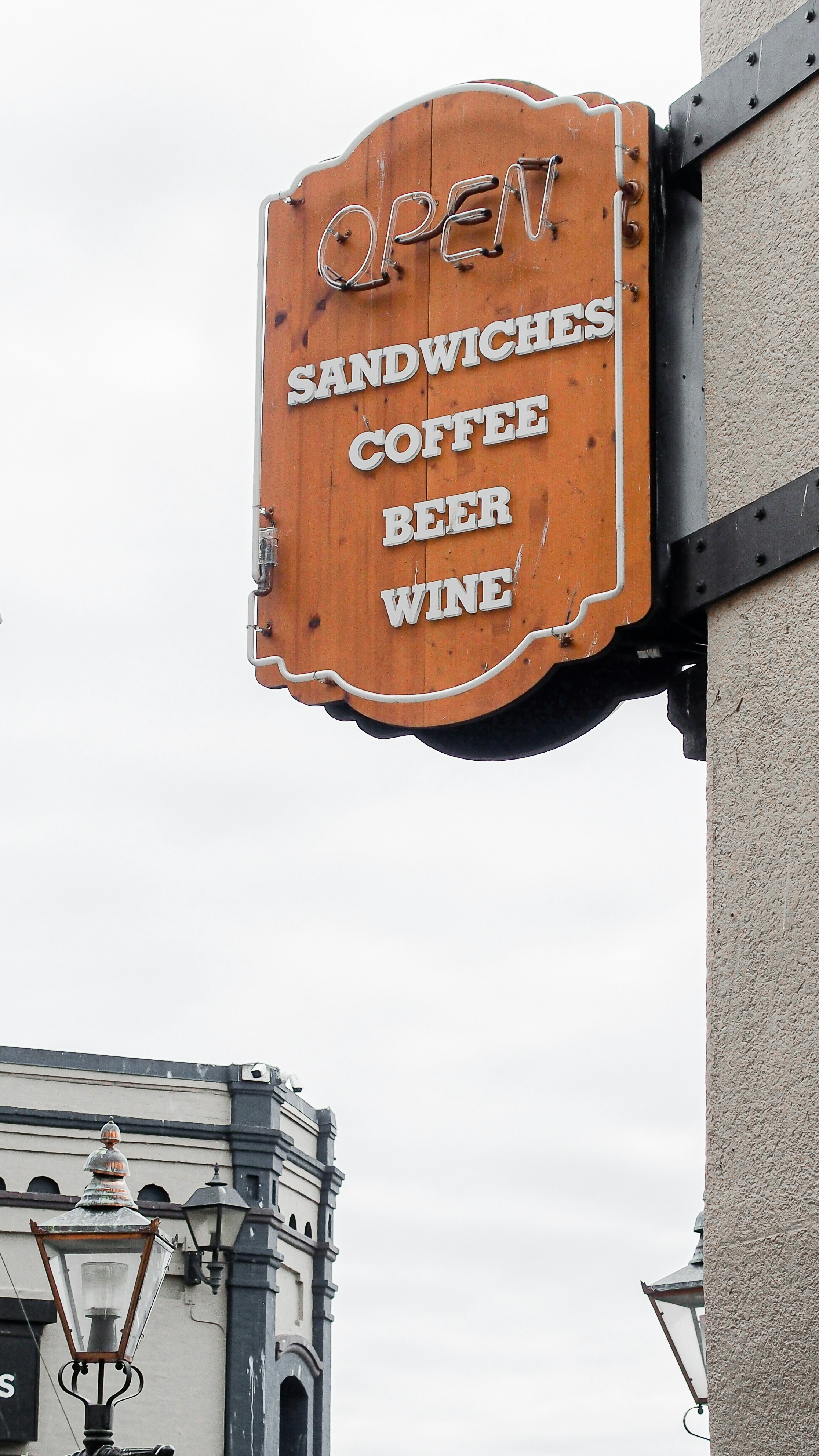 Weathered wooden signboard reading OPEN SANDWICHES COFFEE BEER WINE mounted on the side of a building. The urban storefront photograph captures a rustic cue against a pale sky.