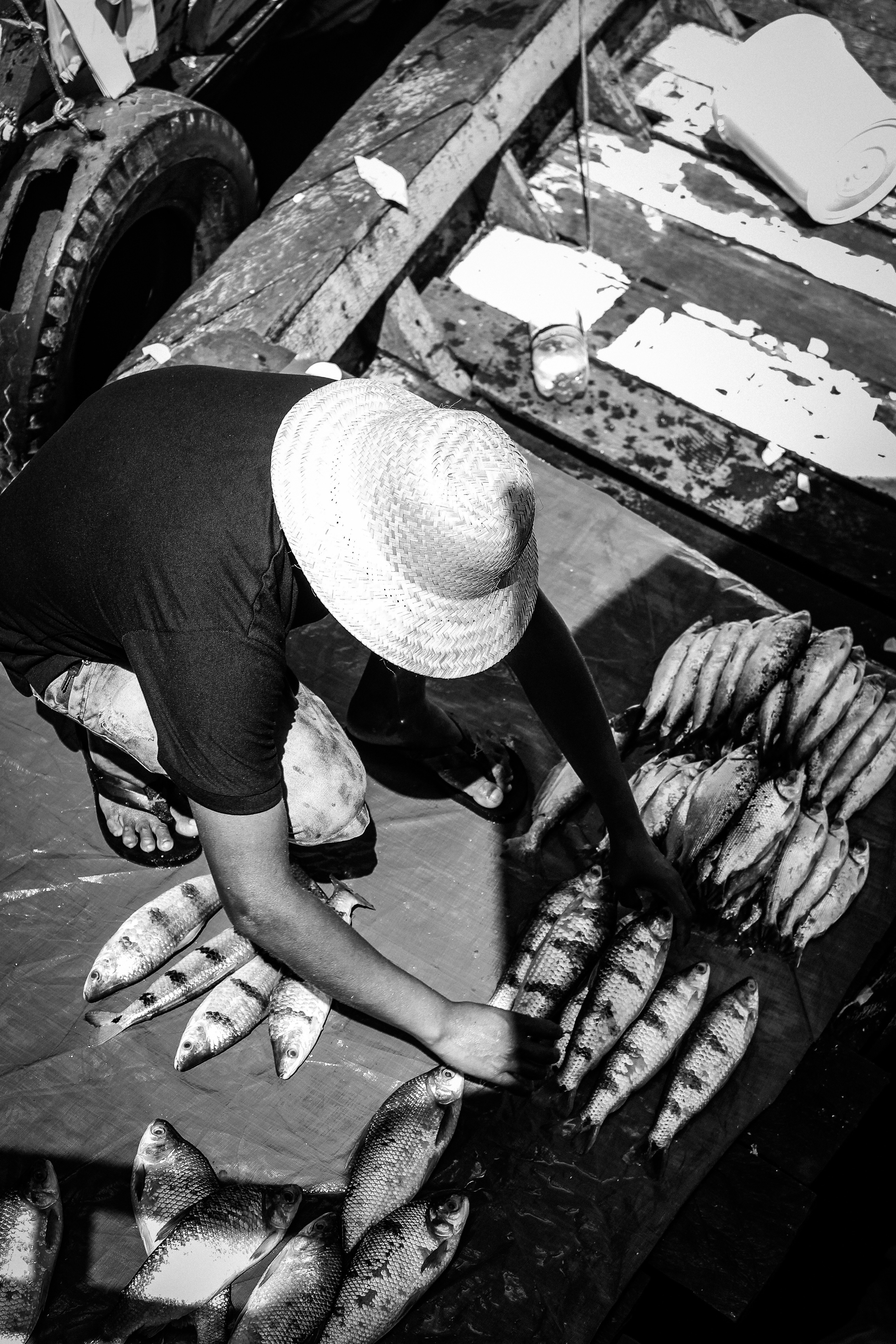 Black-and-white photograph of a worker in a wide-brim hat sorting a pile of fish on a weathered deck.