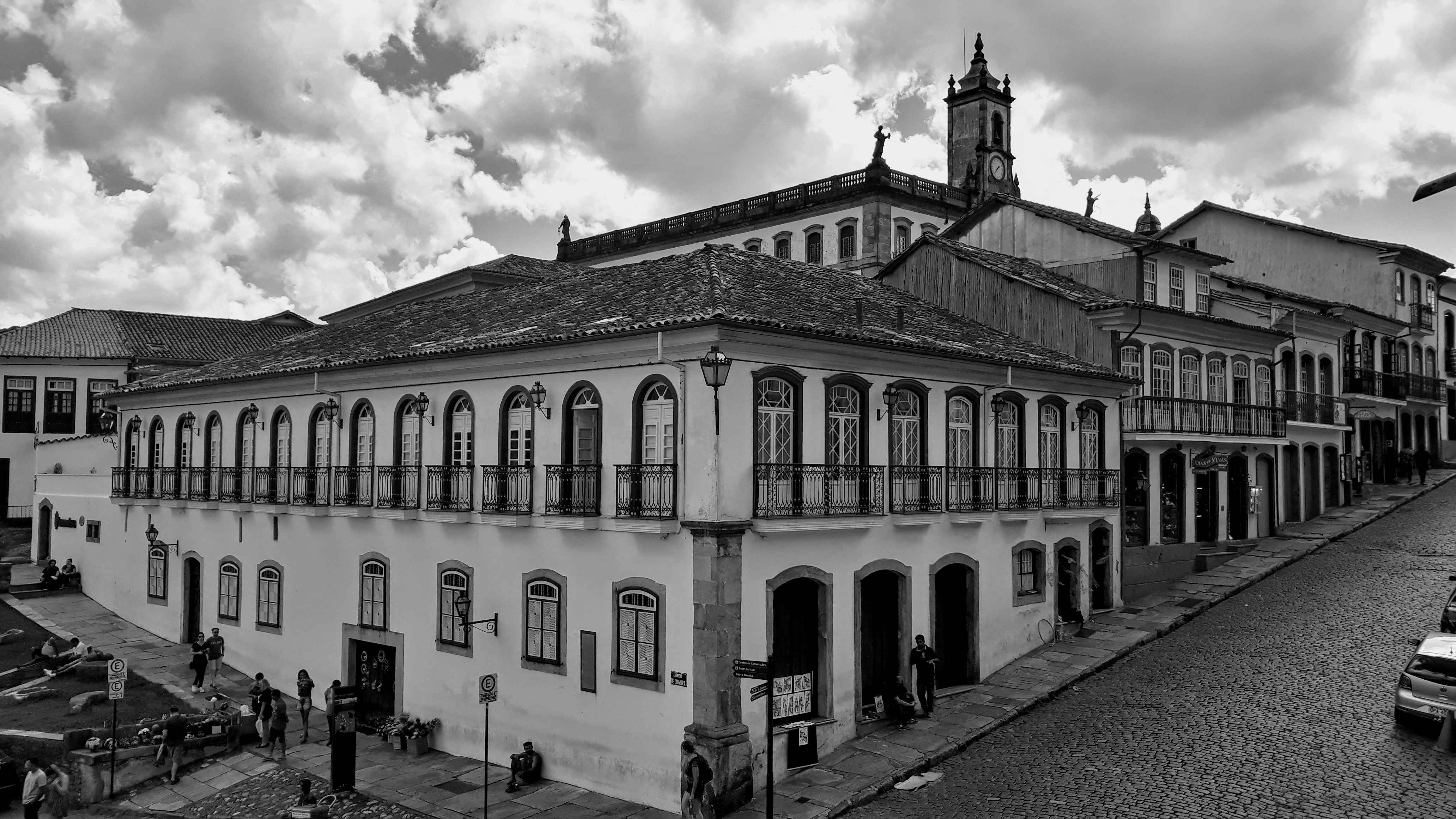 Monochrome view of a historic building with arched windows and a steep street under dramatic clouds.