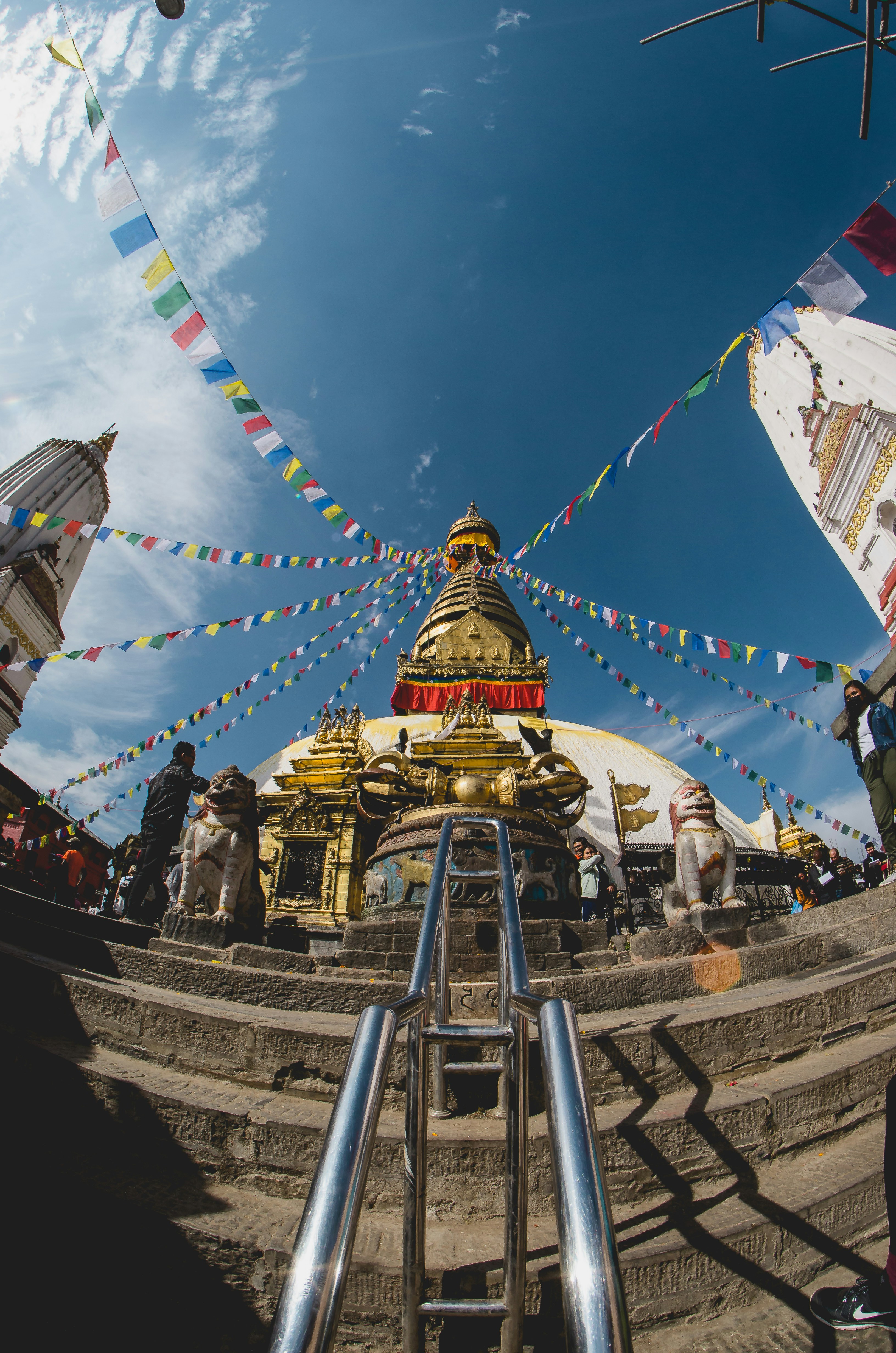 Golden stupa adorned with colorful prayer flags, flanked by lion statues and visitors ascending the steps. Bright blue sky enhances the spiritual ambiance.