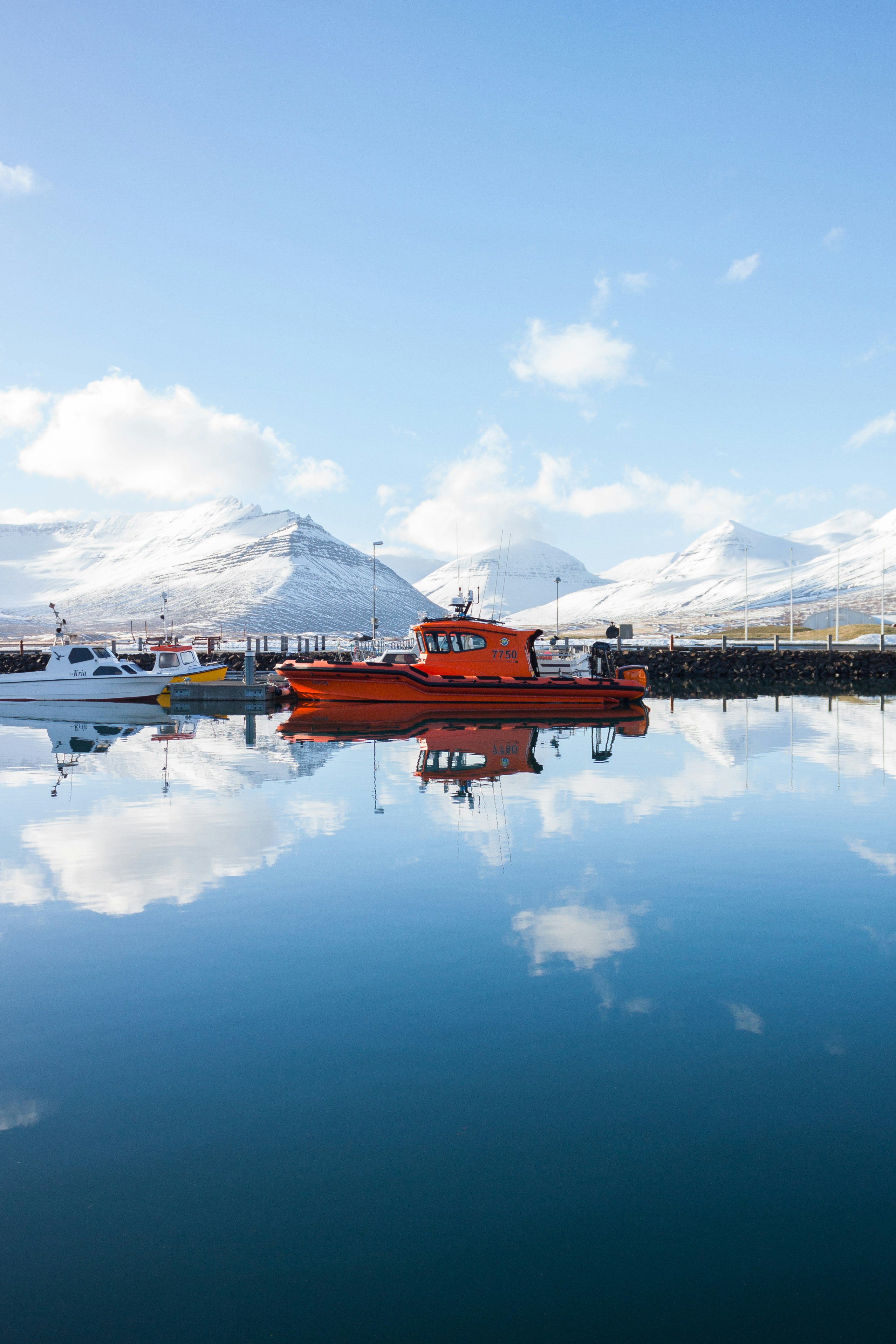 An orange boat anchored in a tranquil harbor, reflecting the snow-capped mountains and blue sky in the still water.