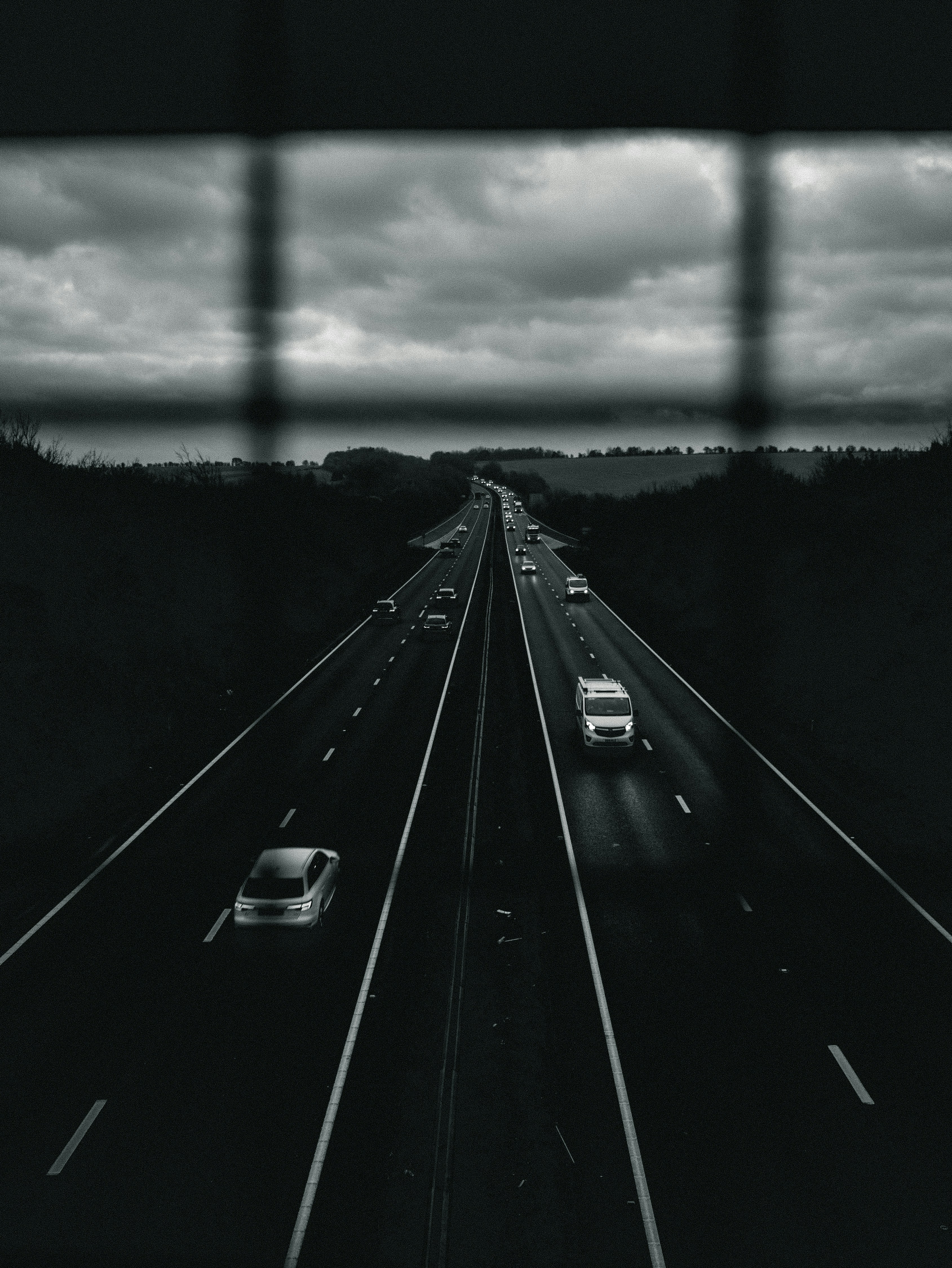 A long stretch of highway under a moody sky, framed by dark silhouettes of trees. Vehicles traverse the road, creating a sense of movement.