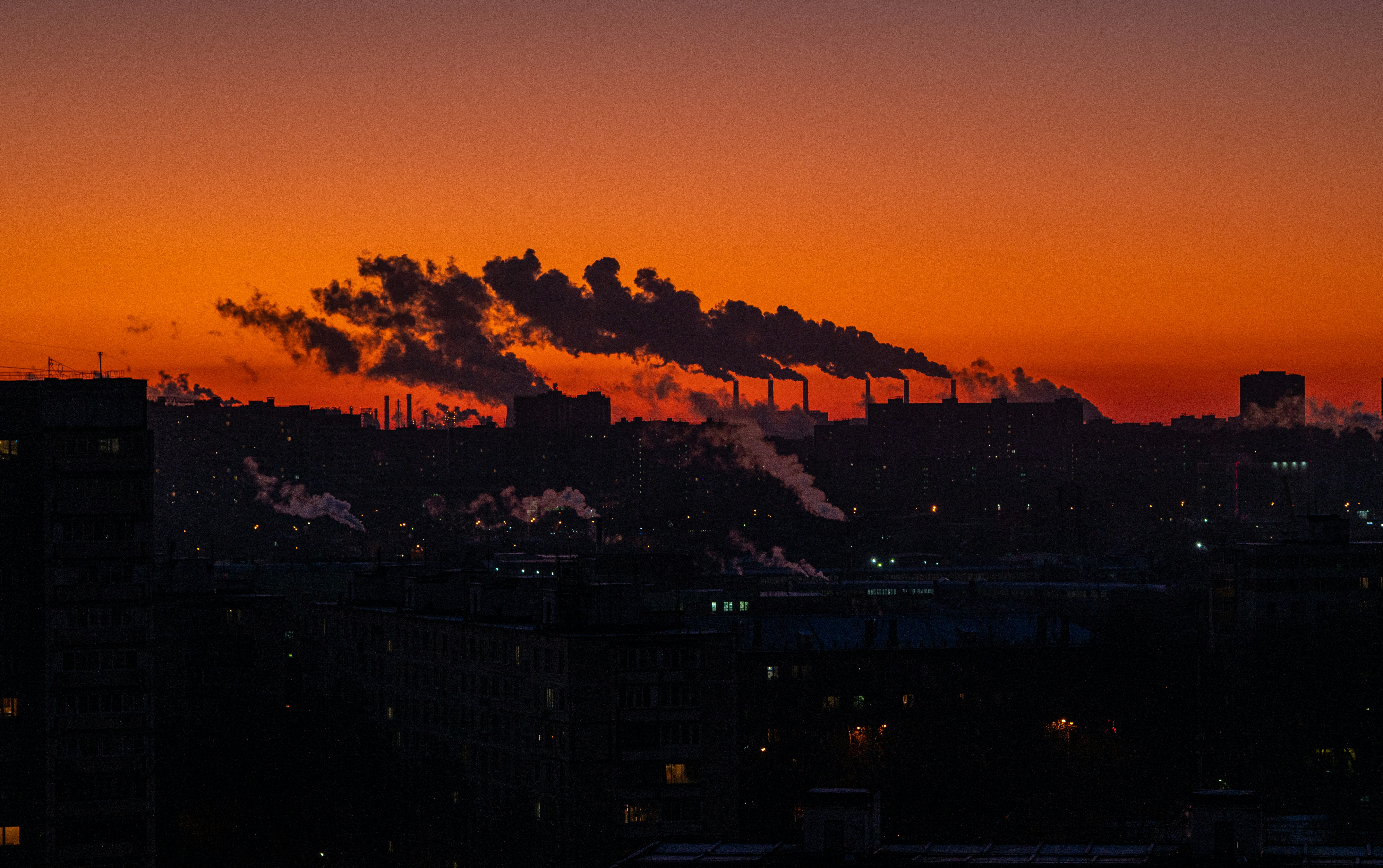 Silhouetted factories emit plumes of smoke against a vibrant sunset, highlighting the contrast between nature and industry.