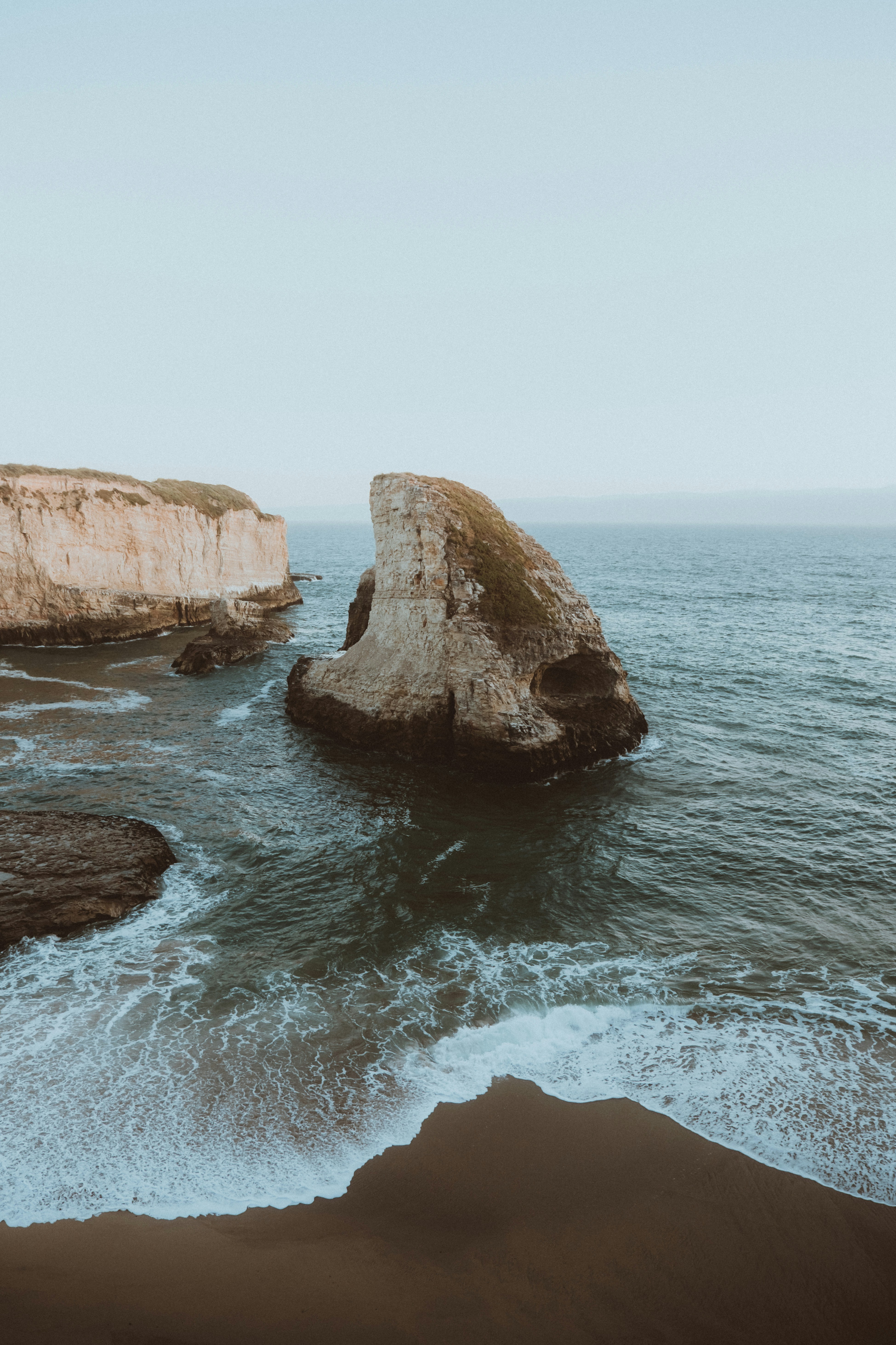 A rugged sea stack rises majestically from the ocean, surrounded by gentle waves and sandy beach. The distant cliffs frame the scene, hinting at the wild beauty of the coast.