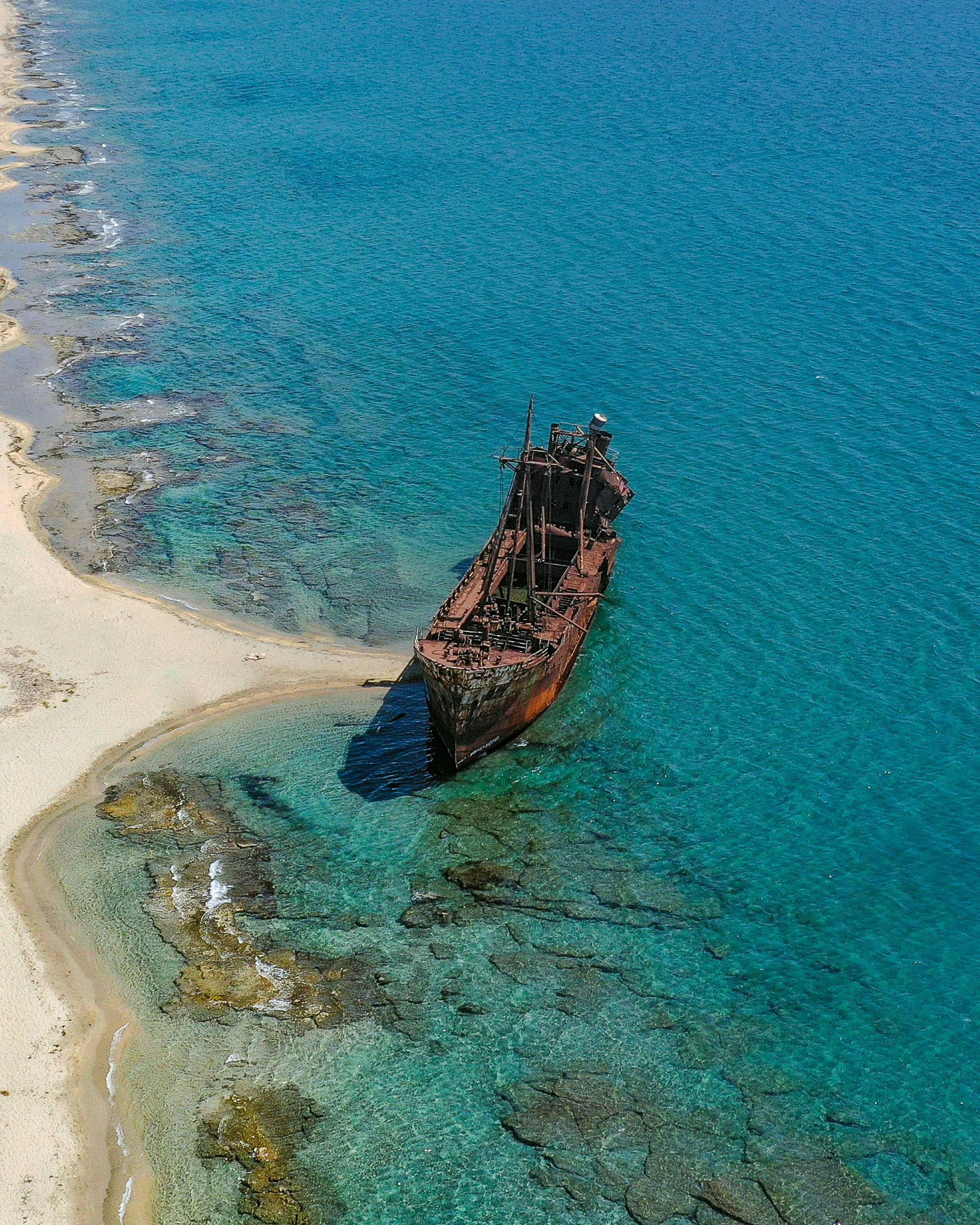 Rusting shipwreck resting on a sandy beach, surrounded by clear turquoise waters and rocky seabed.