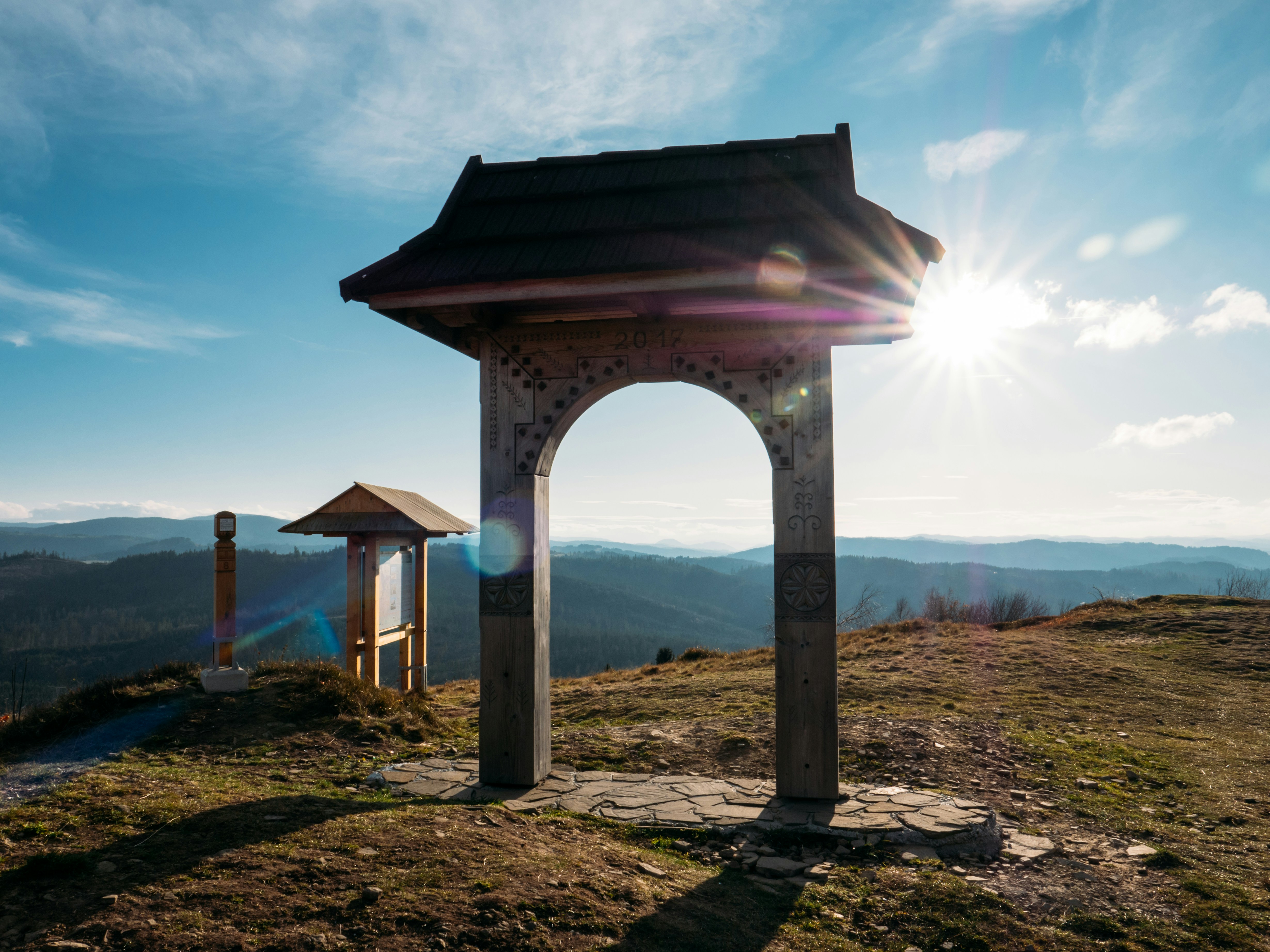 Stone arch on a grassy ridge frames a sunlit valley with distant hills. A small wooden shelter sits to the left, while the bright sun flares behind the arch.