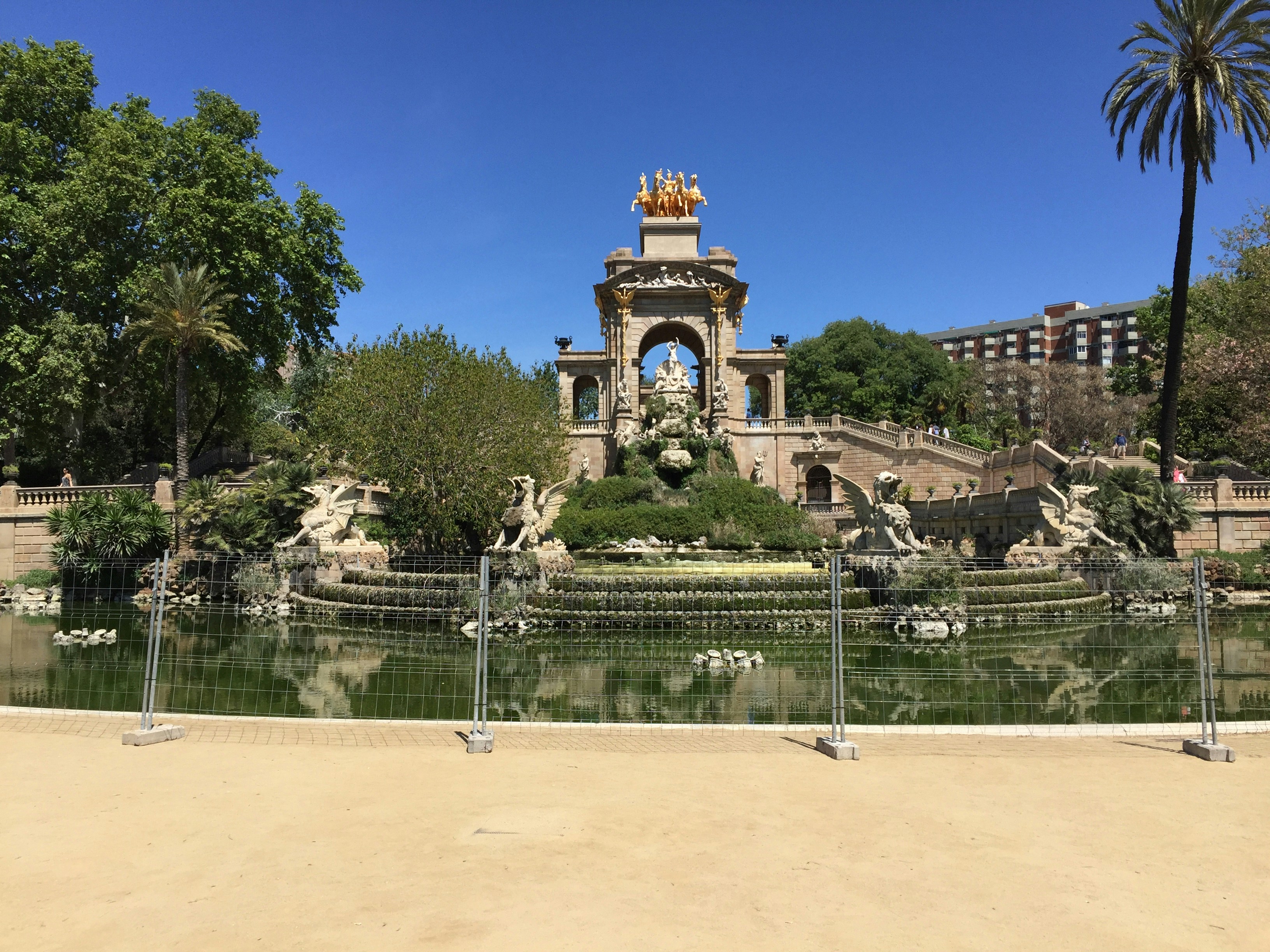 Grand fountain adorned with sculptures surrounded by lush greenery and palm trees in a serene park setting.