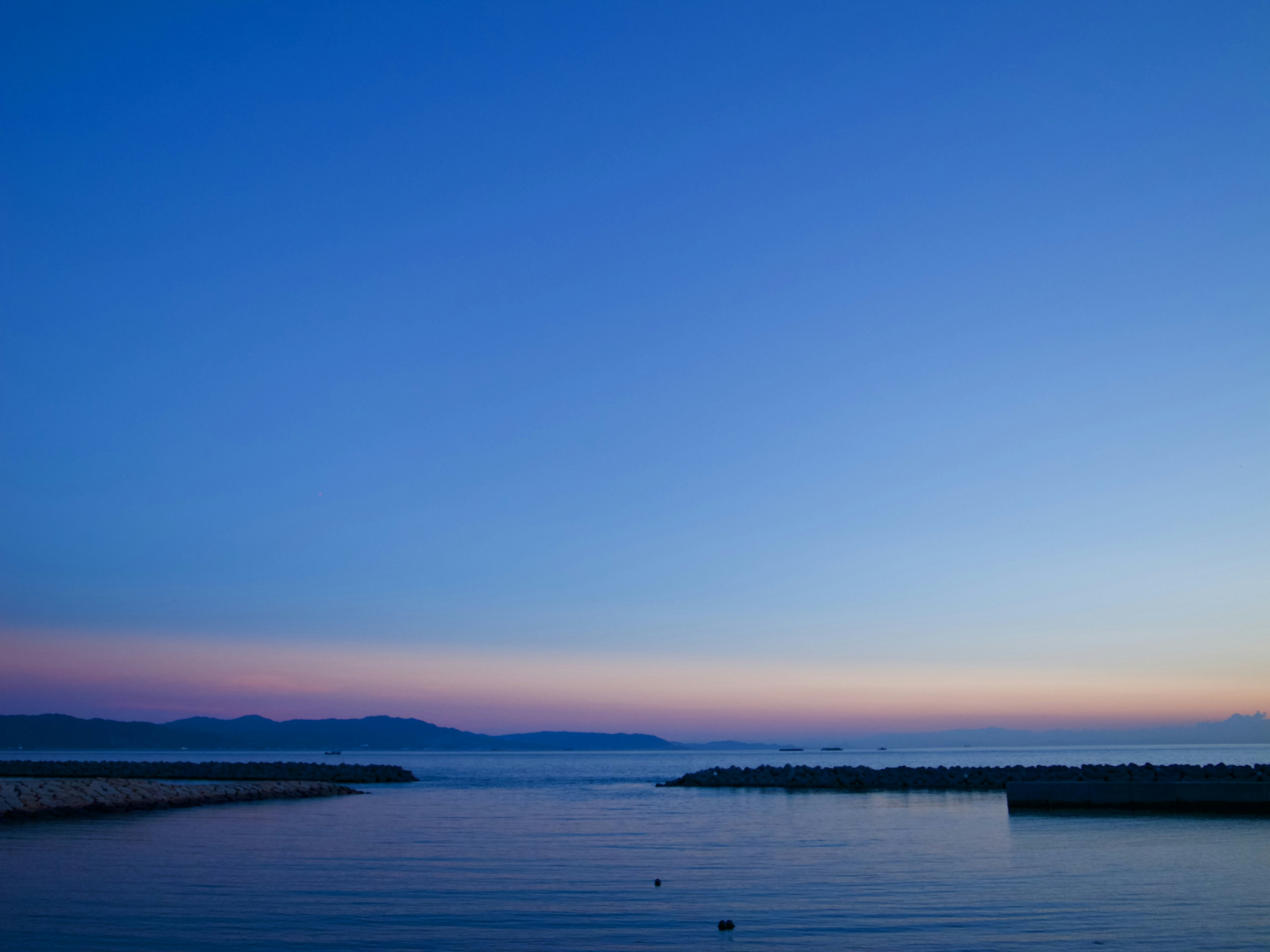 Calm sea at dusk with a pastel pink horizon under a deep blue sky and distant landmasses along the horizon.