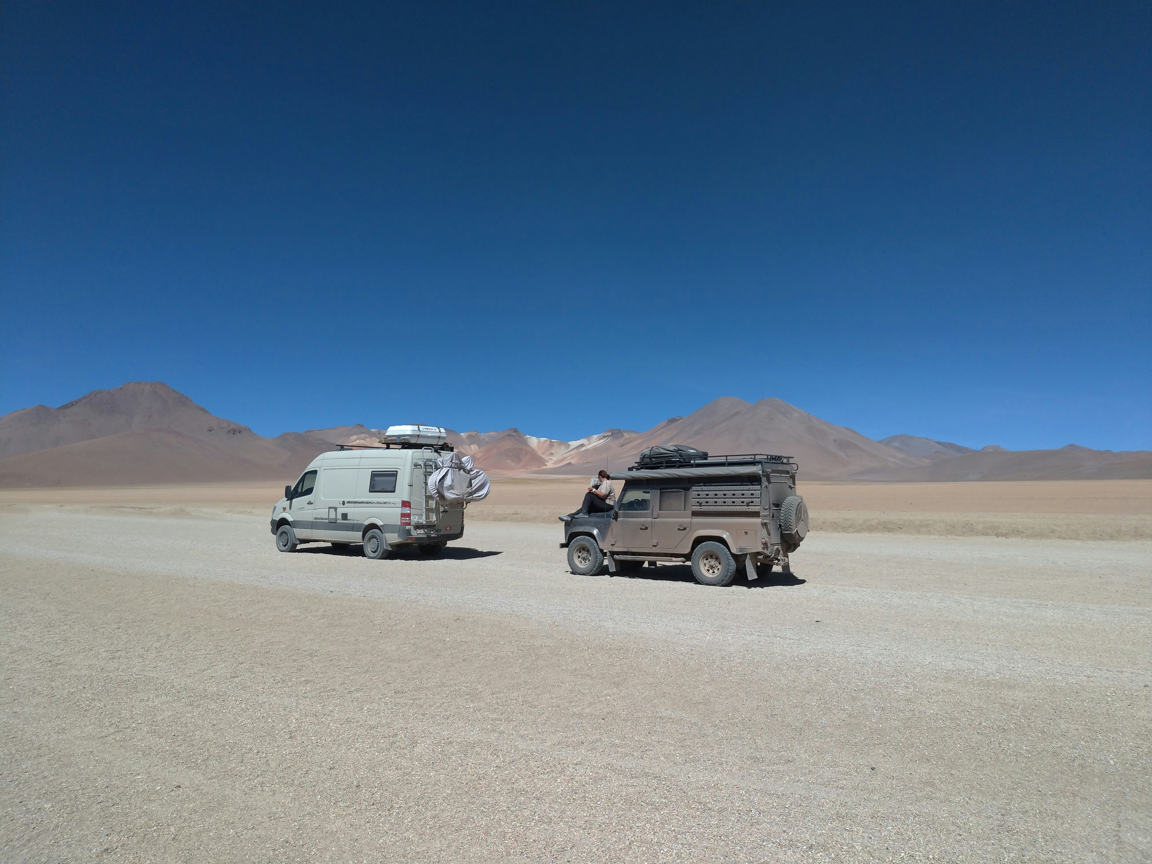 Two rugged vehicles parked on a barren landscape under a clear blue sky, surrounded by distant mountains. The scene captures the essence of adventure and exploration.