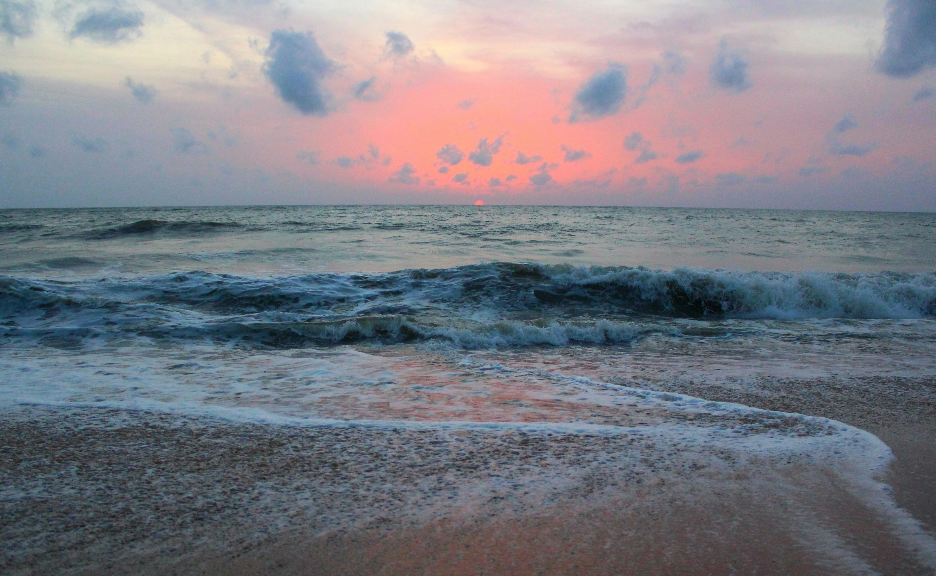 Ocean waves softly crash on a sandy shore under a vibrant pink and purple sunset sky.
