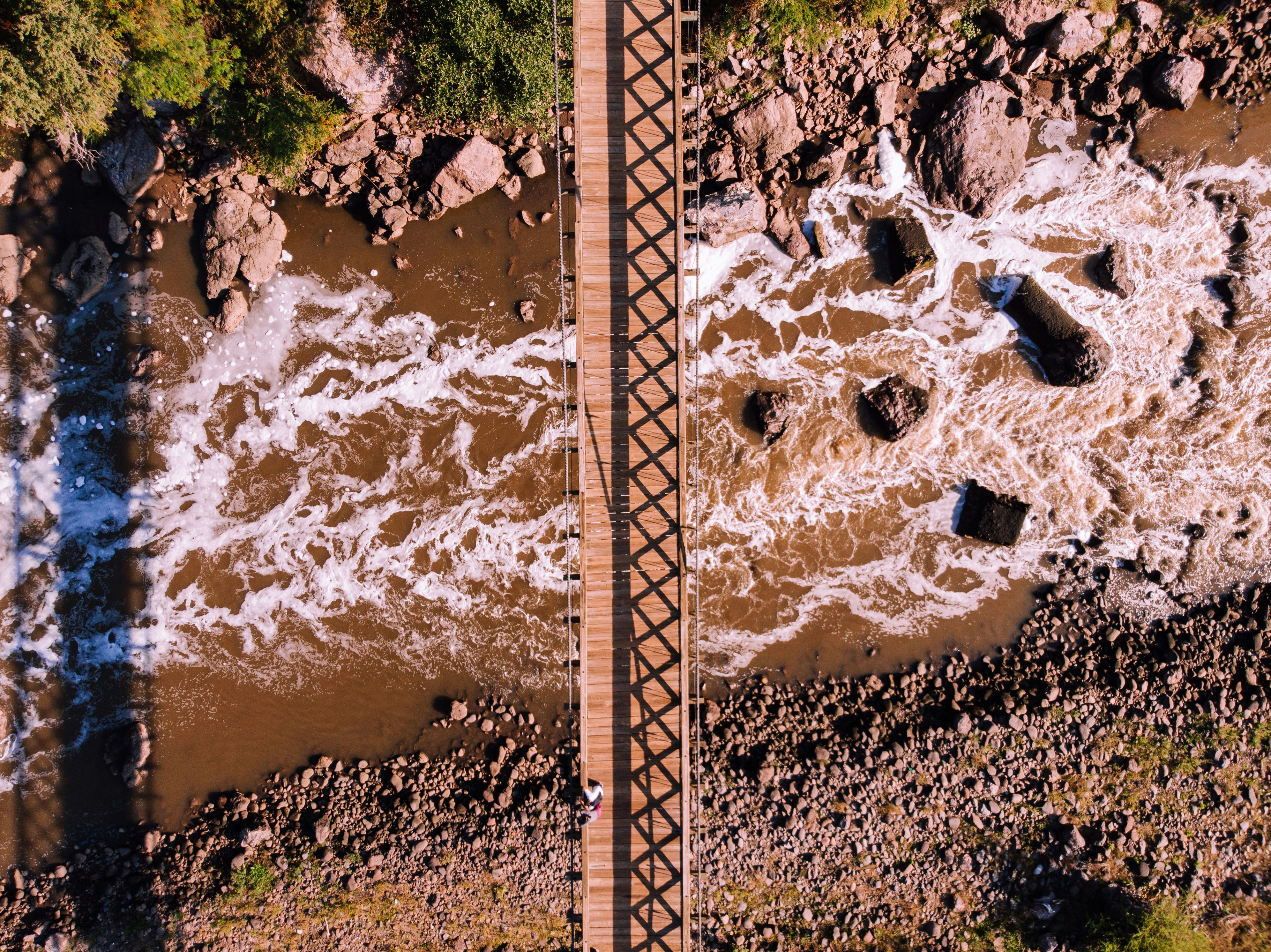 Wooden bridge spanning turbulent river flanked by rocky terrain.