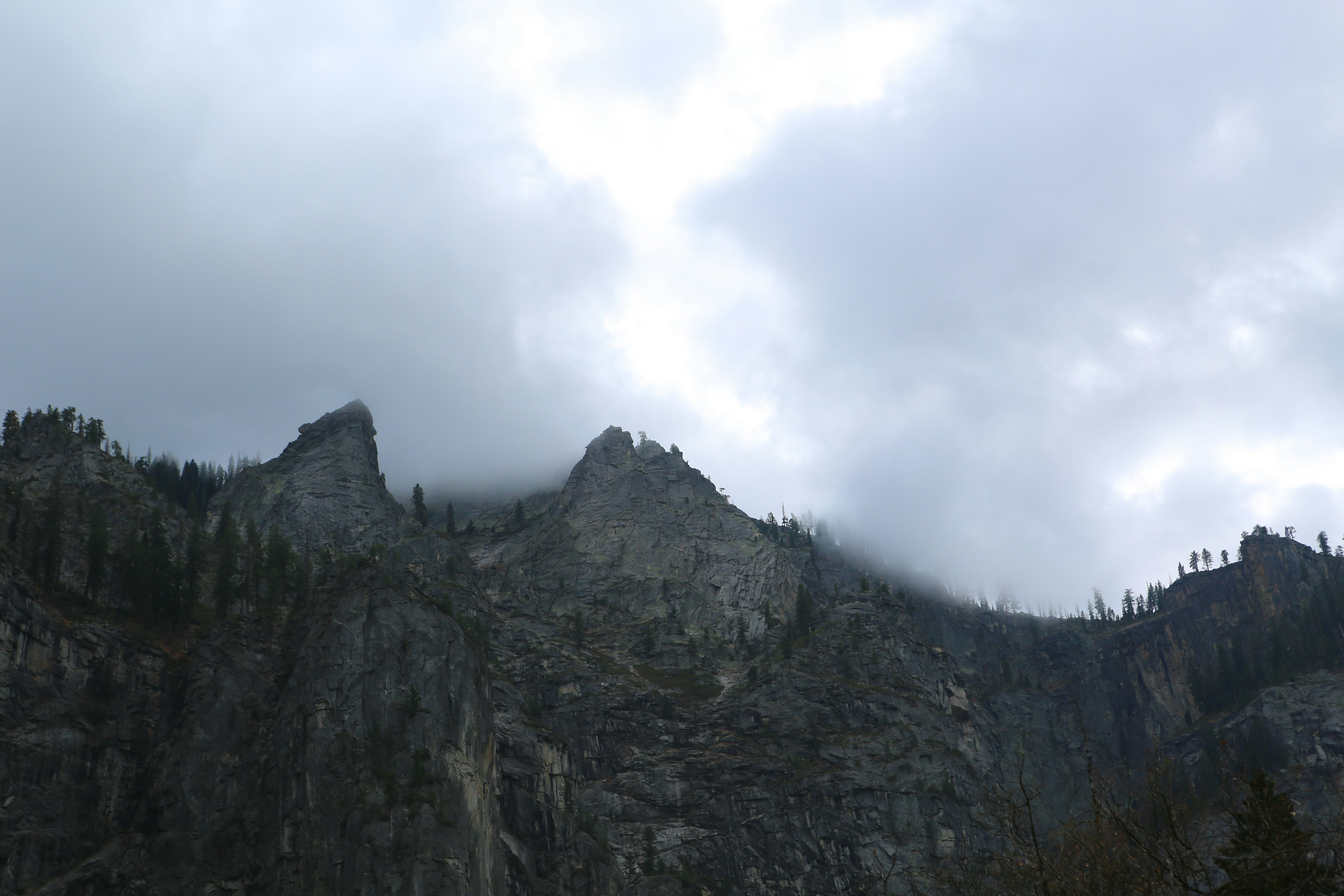 Dramatic mountain peaks partially shrouded in clouds, surrounded by dense forest under a moody sky.