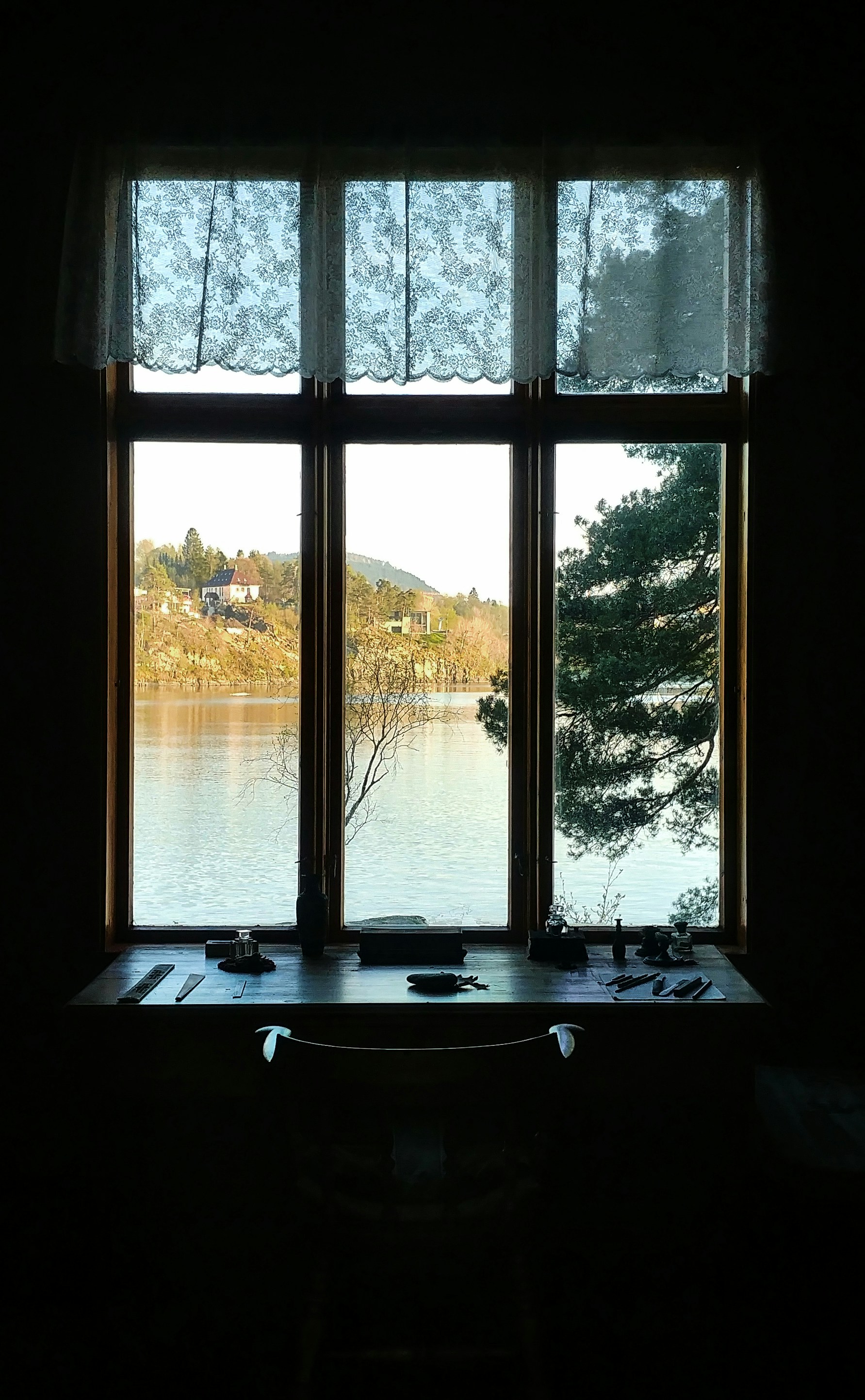 A cozy interior view through a lace-draped window, showcasing a tranquil lake and distant hills. The scene invites reflection and peace.