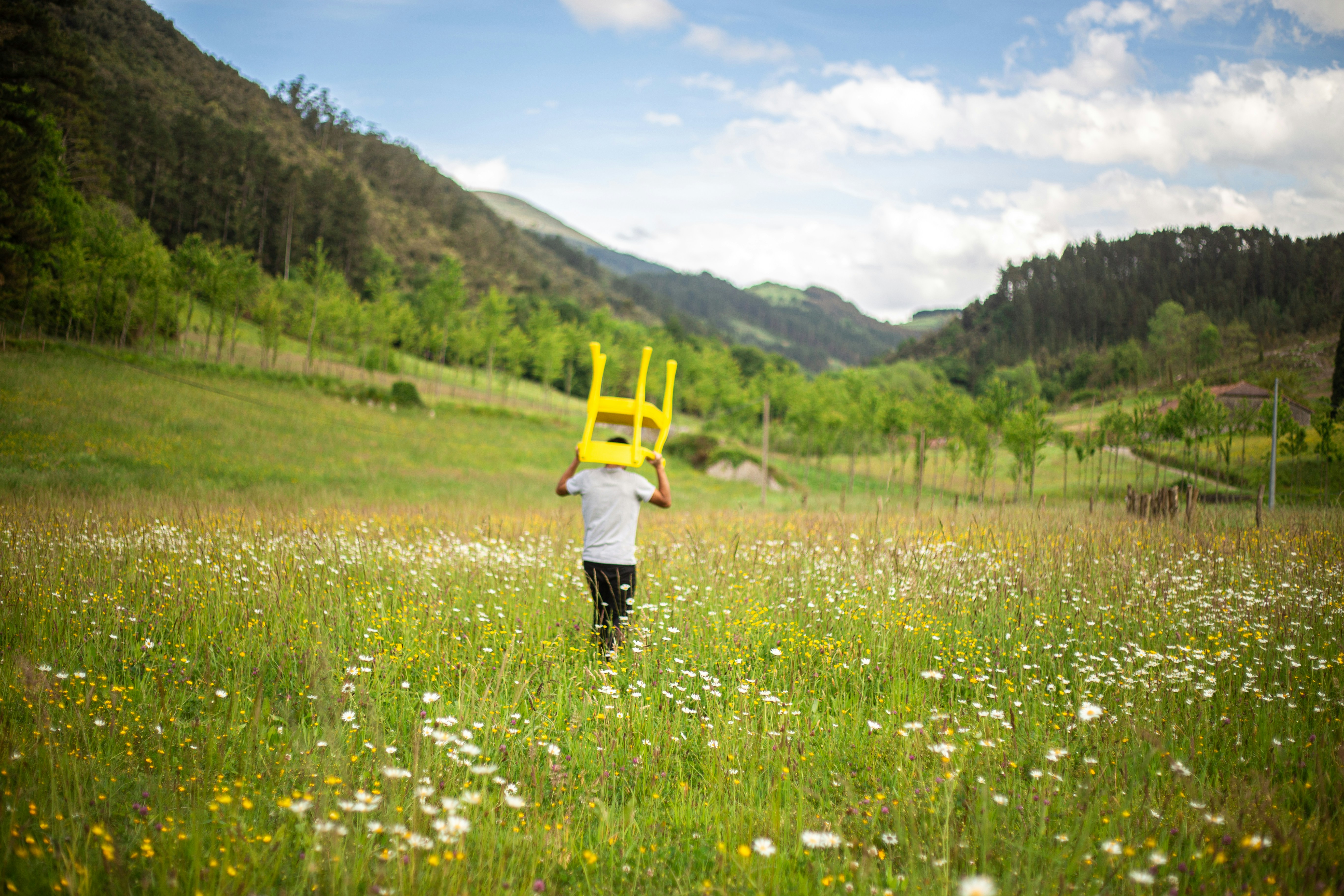 Child carrying a yellow chair across a lush field of wildflowers under a vibrant blue sky.