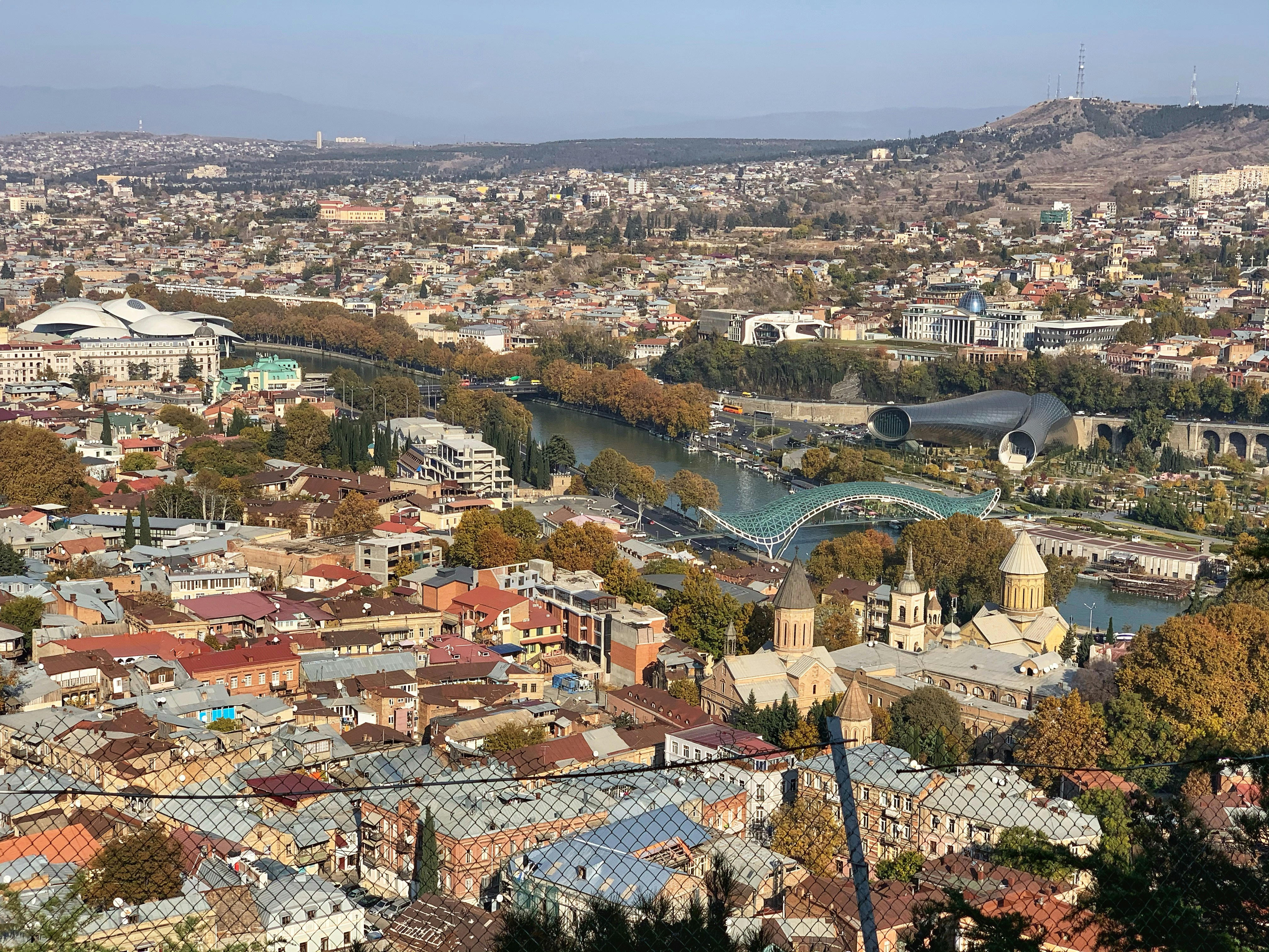Panoramic view of Tbilisi showcasing a blend of modern architecture and historic structures along the river. Autumn foliage adds warmth to the urban landscape.