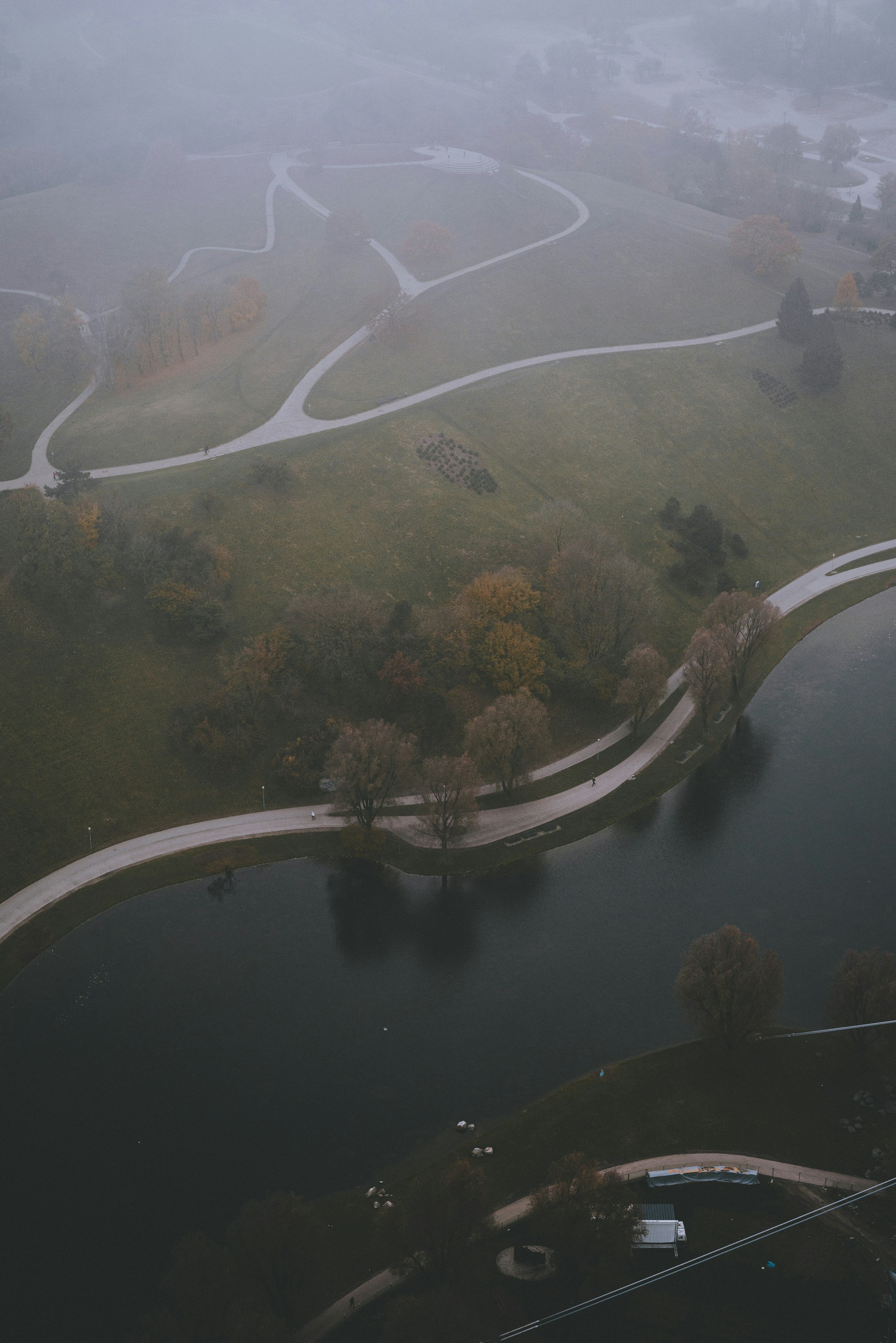 Aerial view of winding paths and a tranquil lake surrounded by trees in a foggy landscape. The soft hues create a peaceful atmosphere.