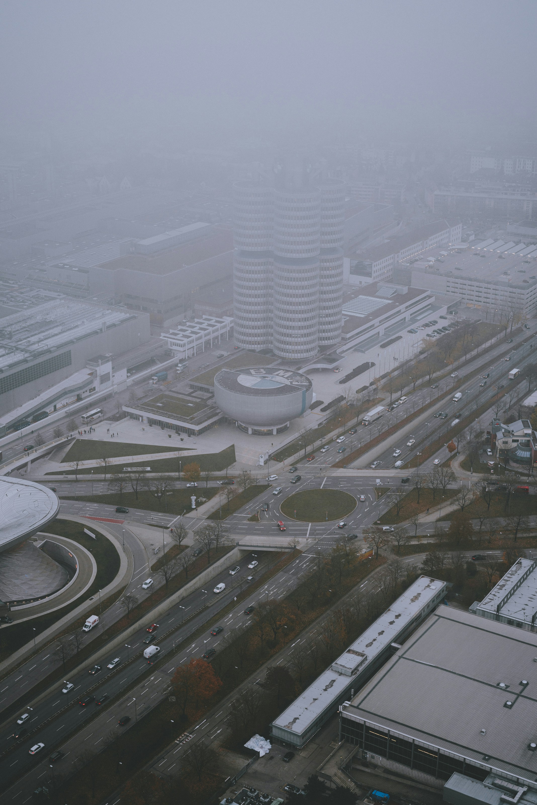 Aerial view of a modern urban landscape shrouded in fog, featuring a prominent circular building and winding roads below.