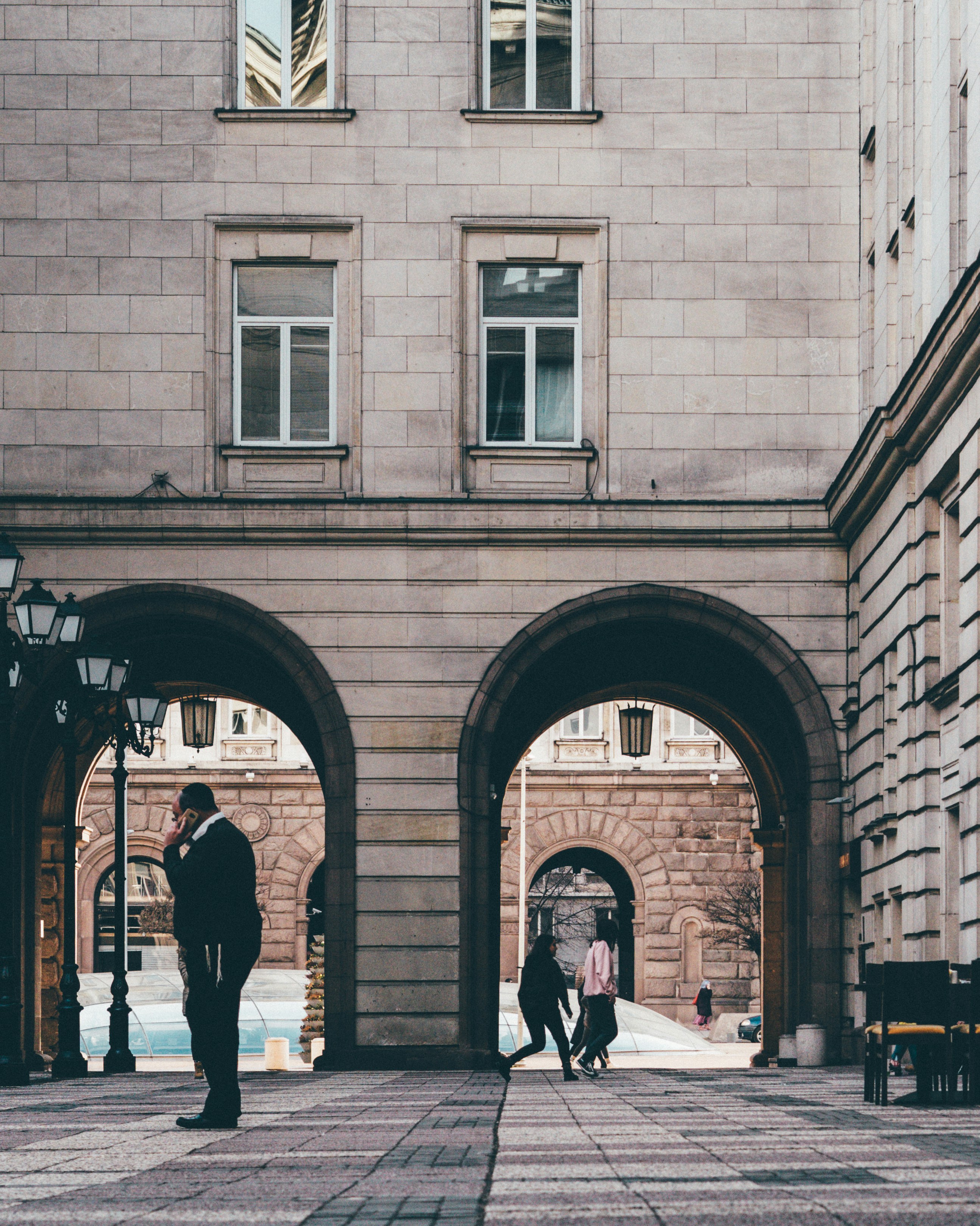 People walking through an archway in a historic urban setting, framed by elegant architecture and street lamps.