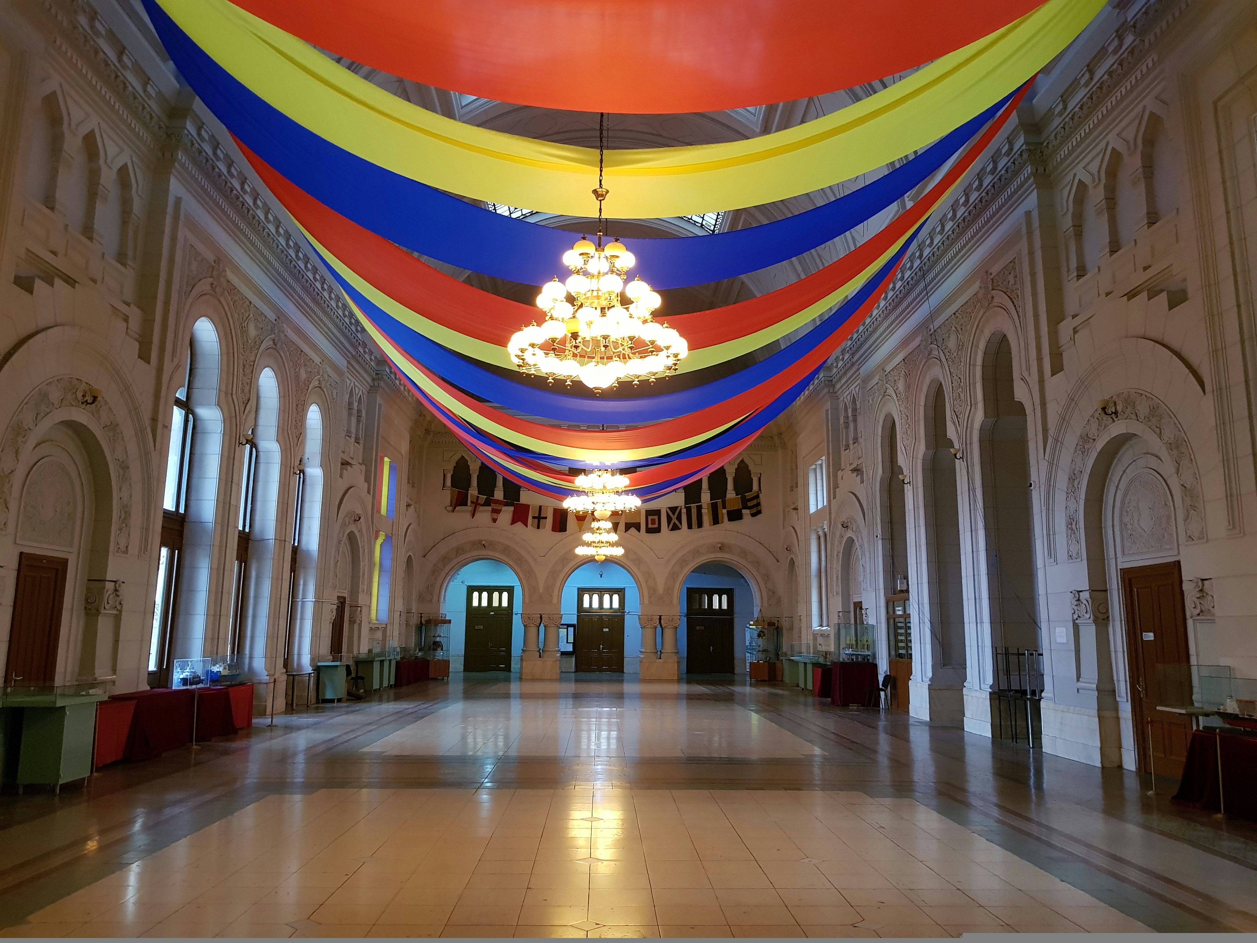 Colorful drapes hang beneath ornate chandeliers in an elegant hall with arched windows.