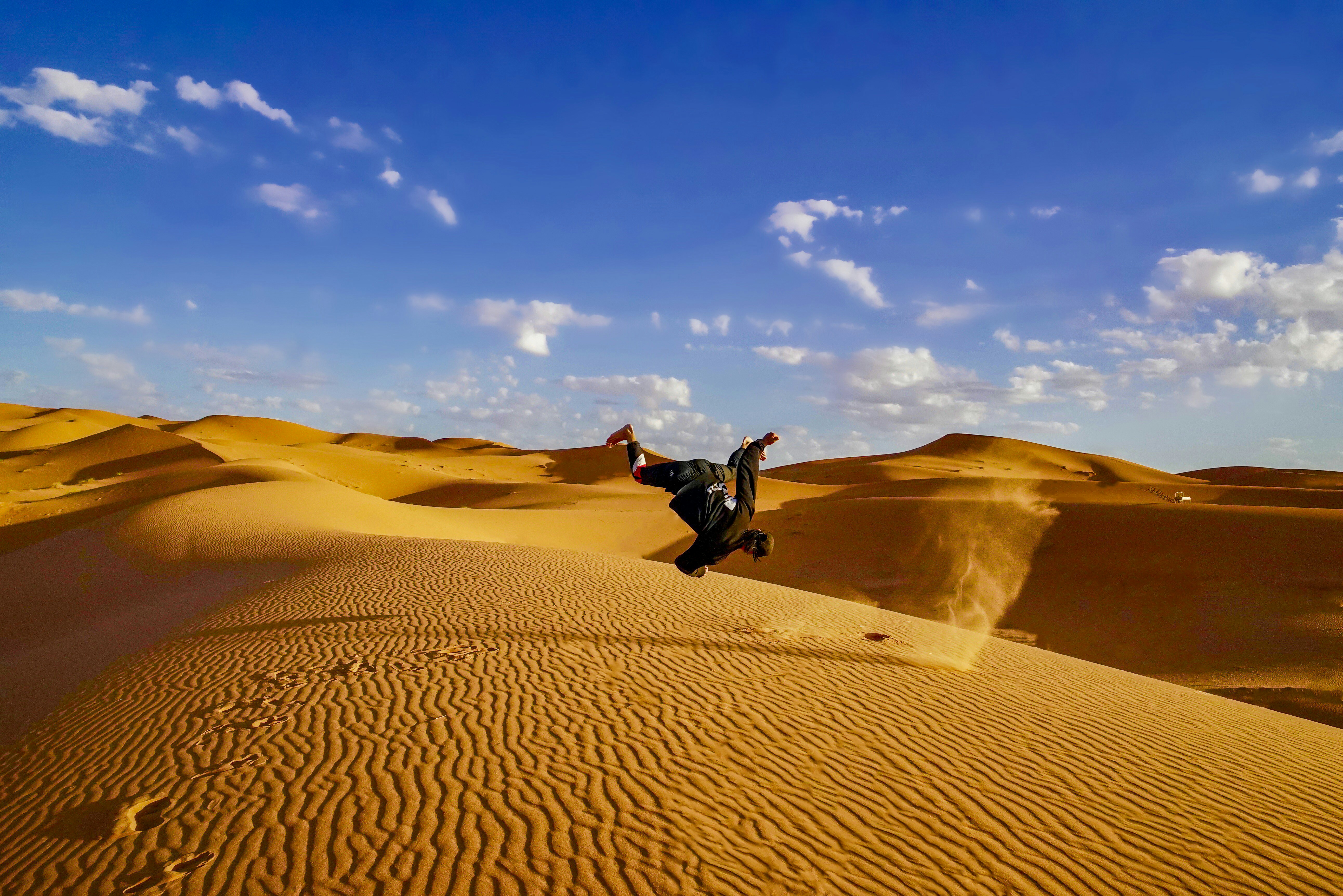 A figure leaps gracefully across the undulating dunes of a vast desert, kicking up sand in a dynamic display of movement. The scene captures the essence of freedom and adventure in a striking landscape.