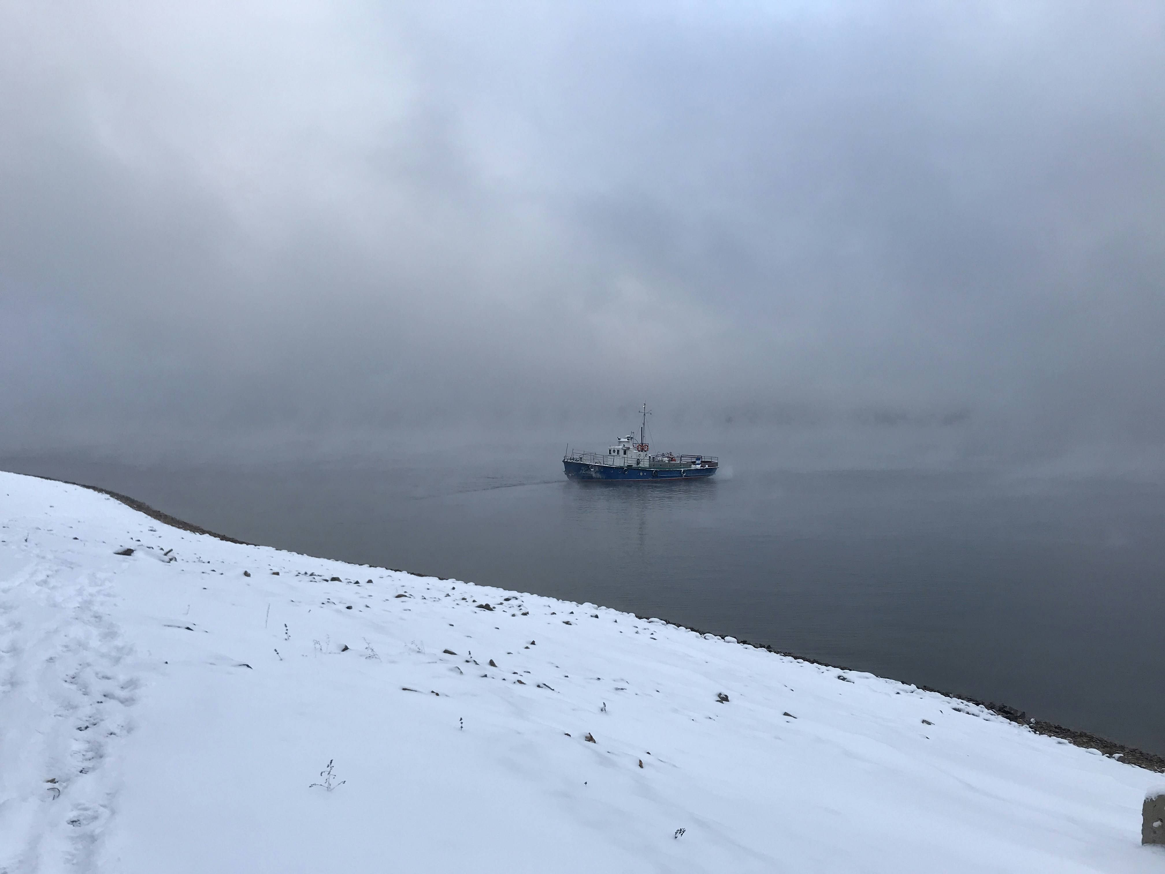 A solitary boat glides across a calm, foggy river, framed by a snowy shoreline. The scene evokes a sense of tranquility and solitude.