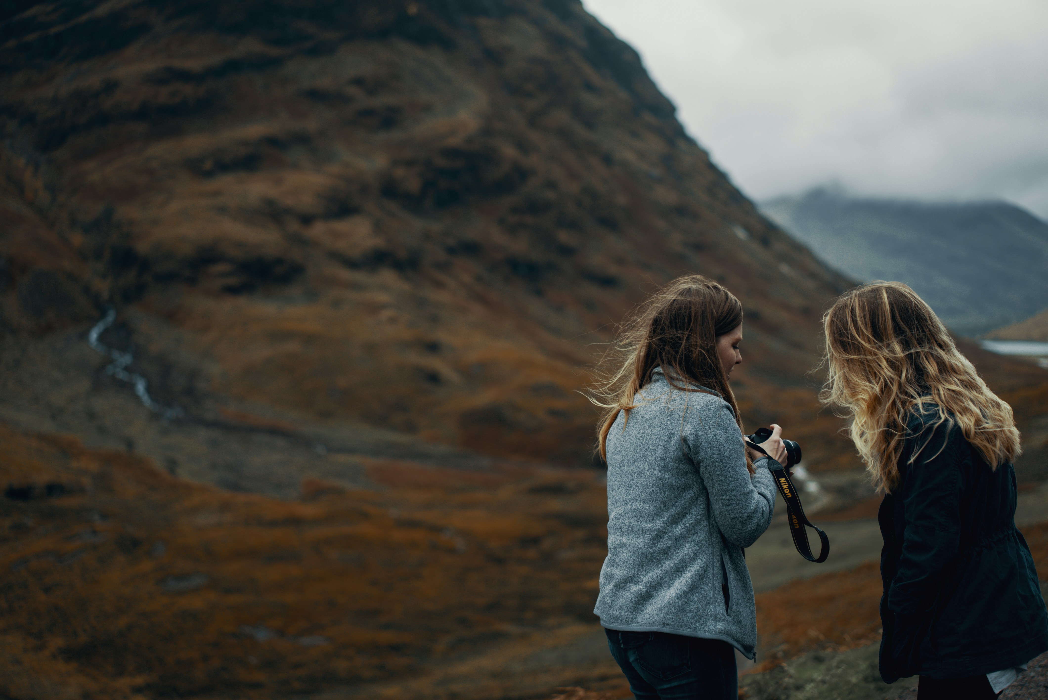 Two friends with a camera stand on a hillside, preparing for a shot against a moody autumn landscape with brown slopes and distant mountains.