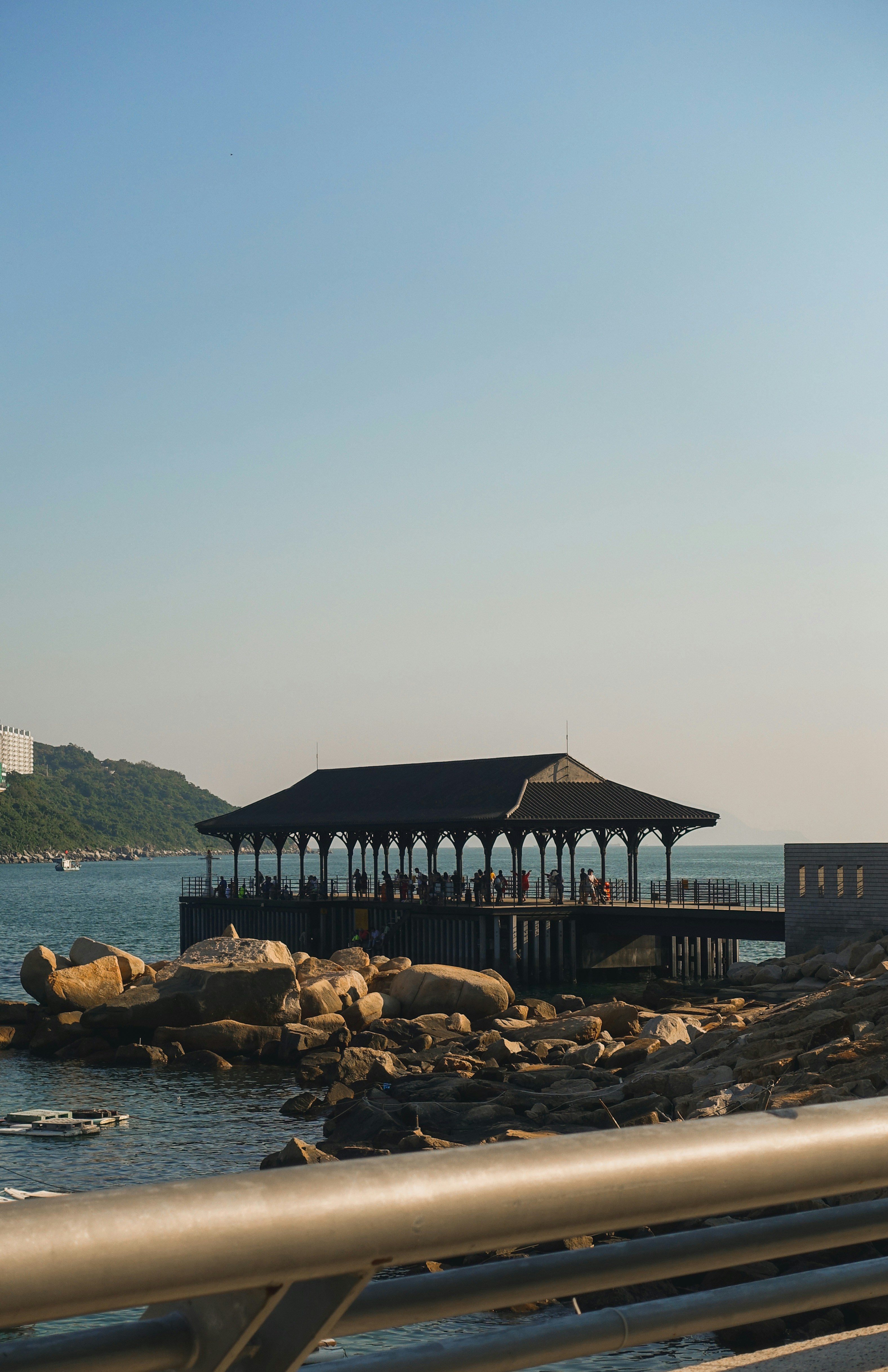 A wooden pavilion perched over rocky shores, inviting visitors to enjoy the serene coastal views. The calm water reflects the clear sky.