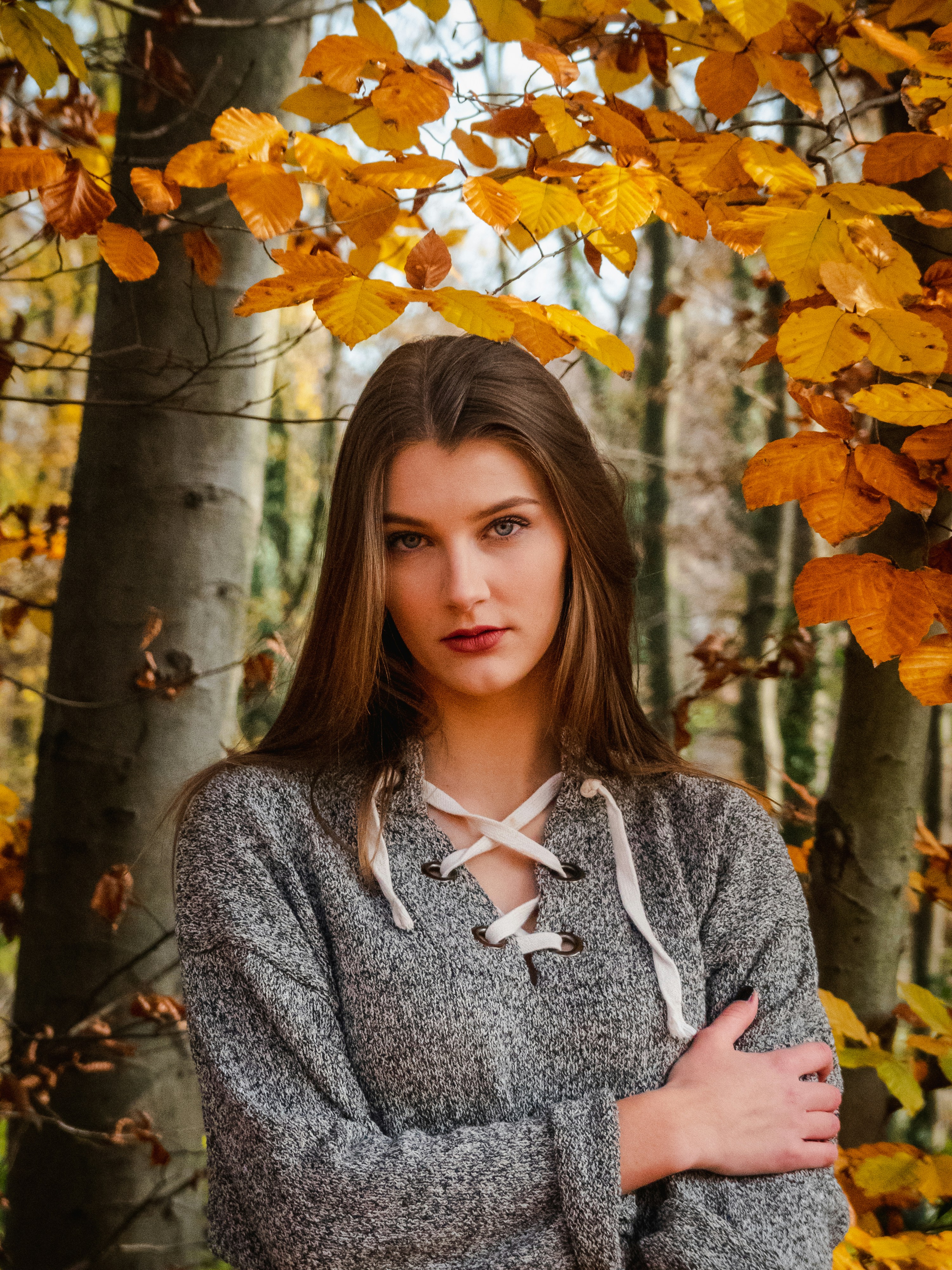 A young woman stands gracefully amidst vibrant autumn foliage, her expression reflecting a serene confidence. The warm tones of the leaves create a striking contrast with her attire.