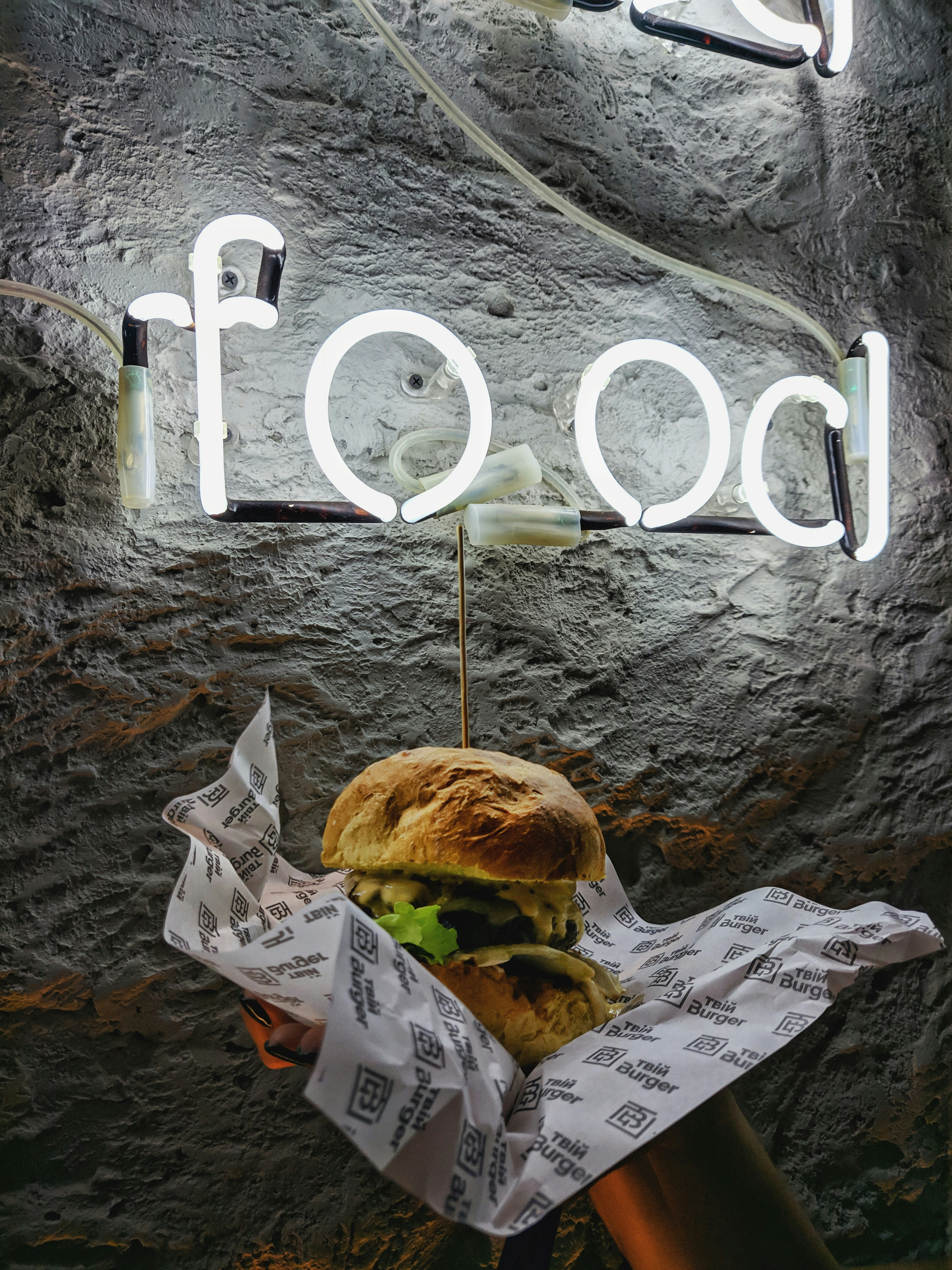 A freshly made burger held in hand, wrapped in branded paper, with a glowing neon sign that reads 'food' in the background.