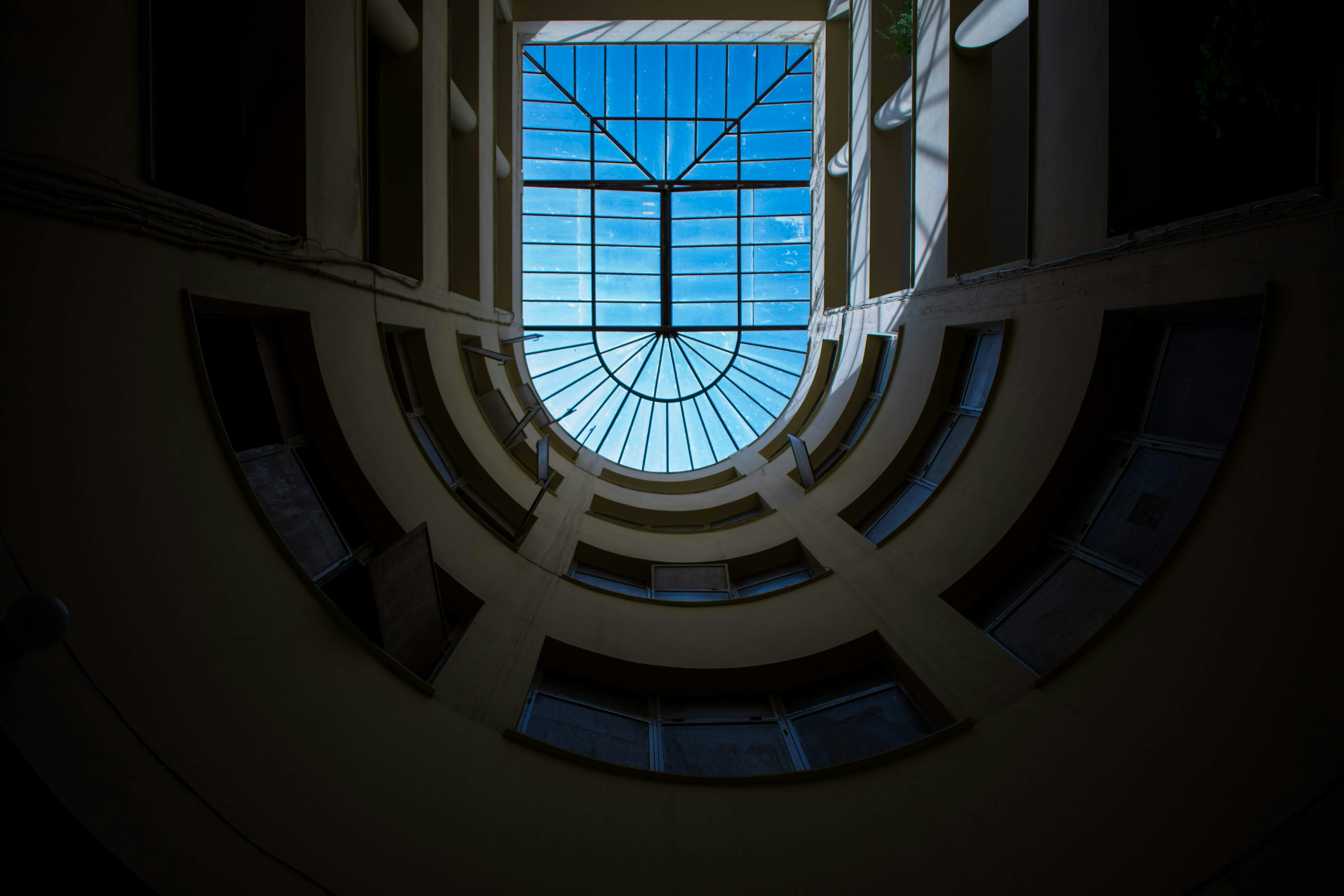 Interior view of a circular building, showcasing a skylight framed by curved walls and windows. Bright blue sky contrasts with the surrounding structure.