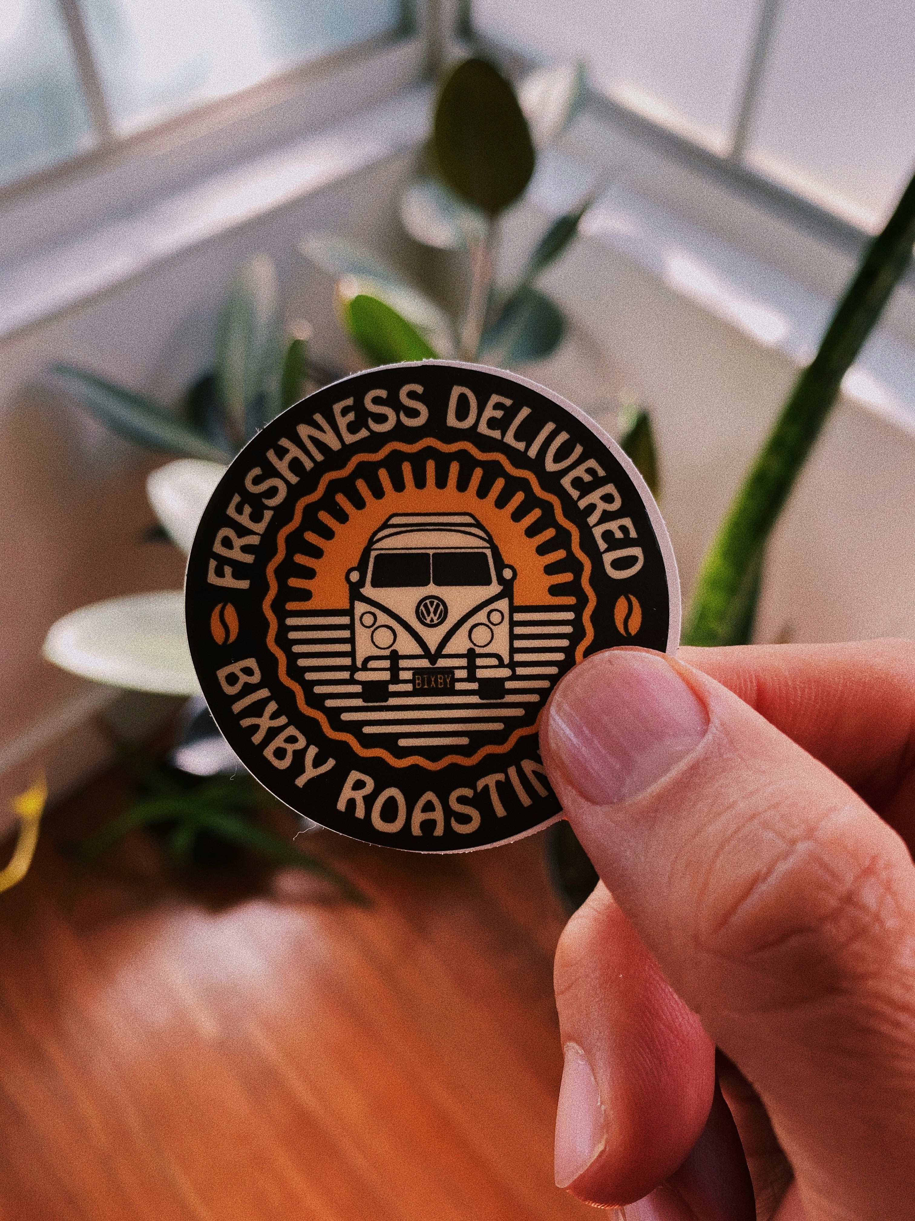 Round logo-style sticker featuring a vintage Volkswagen bus and the text 'Freshness Delivered'. It is held between fingers with indoor plants and a window blurred in the background.