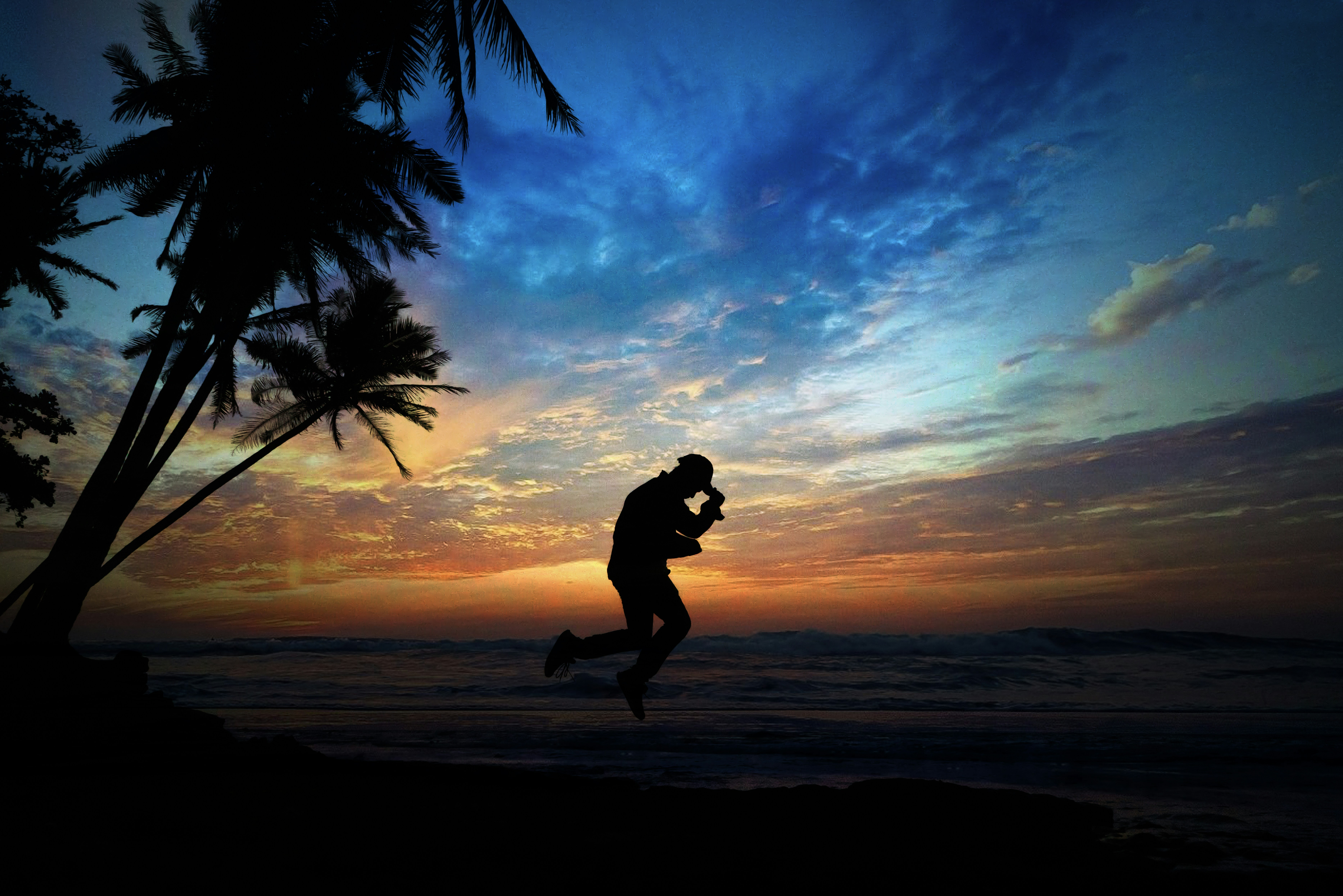Silhouette of a person photographing a vibrant sunrise on a beach framed by palm trees.