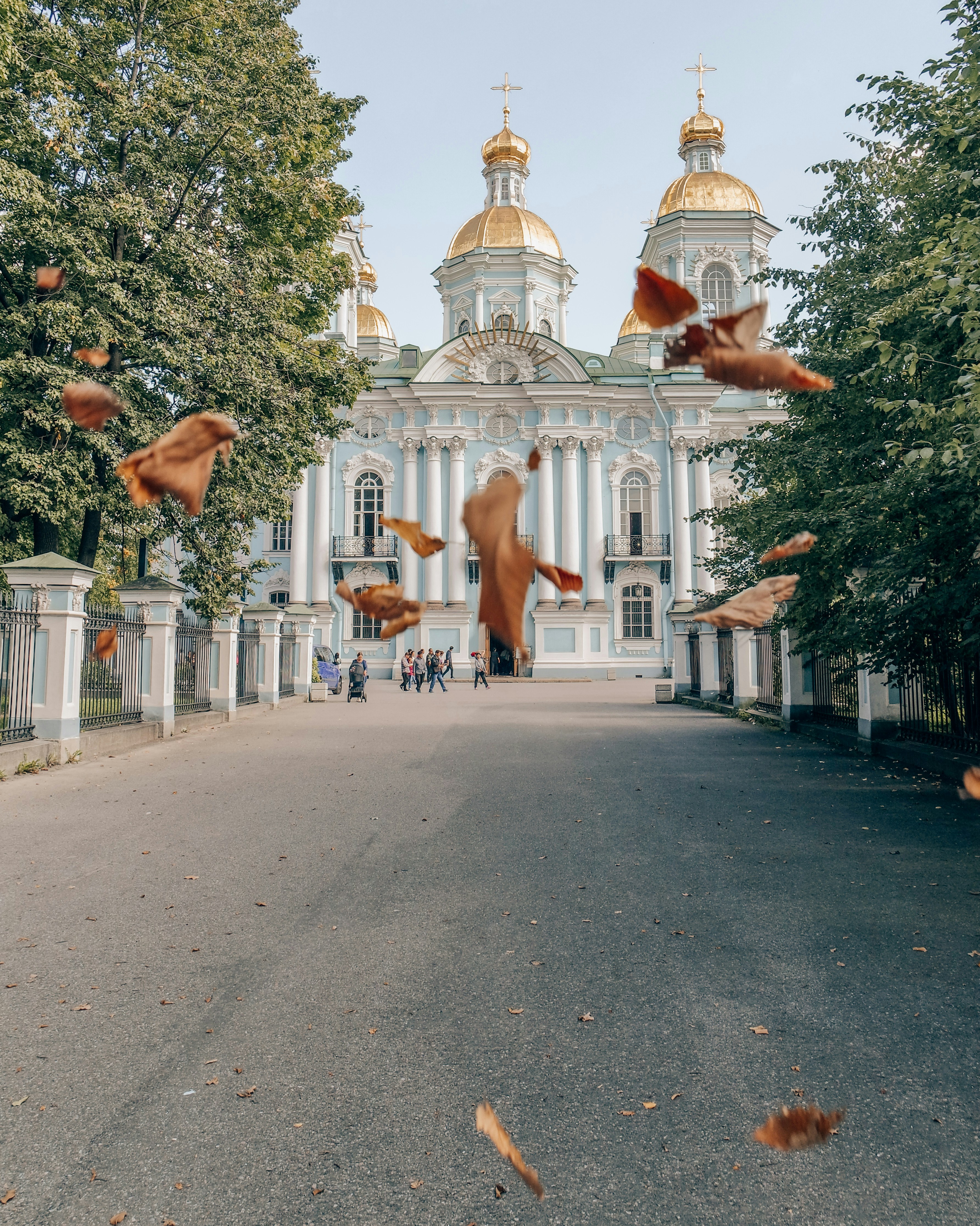 Golden leaves dance in the air near a grand cathedral, framed by lush greenery and a serene pathway.