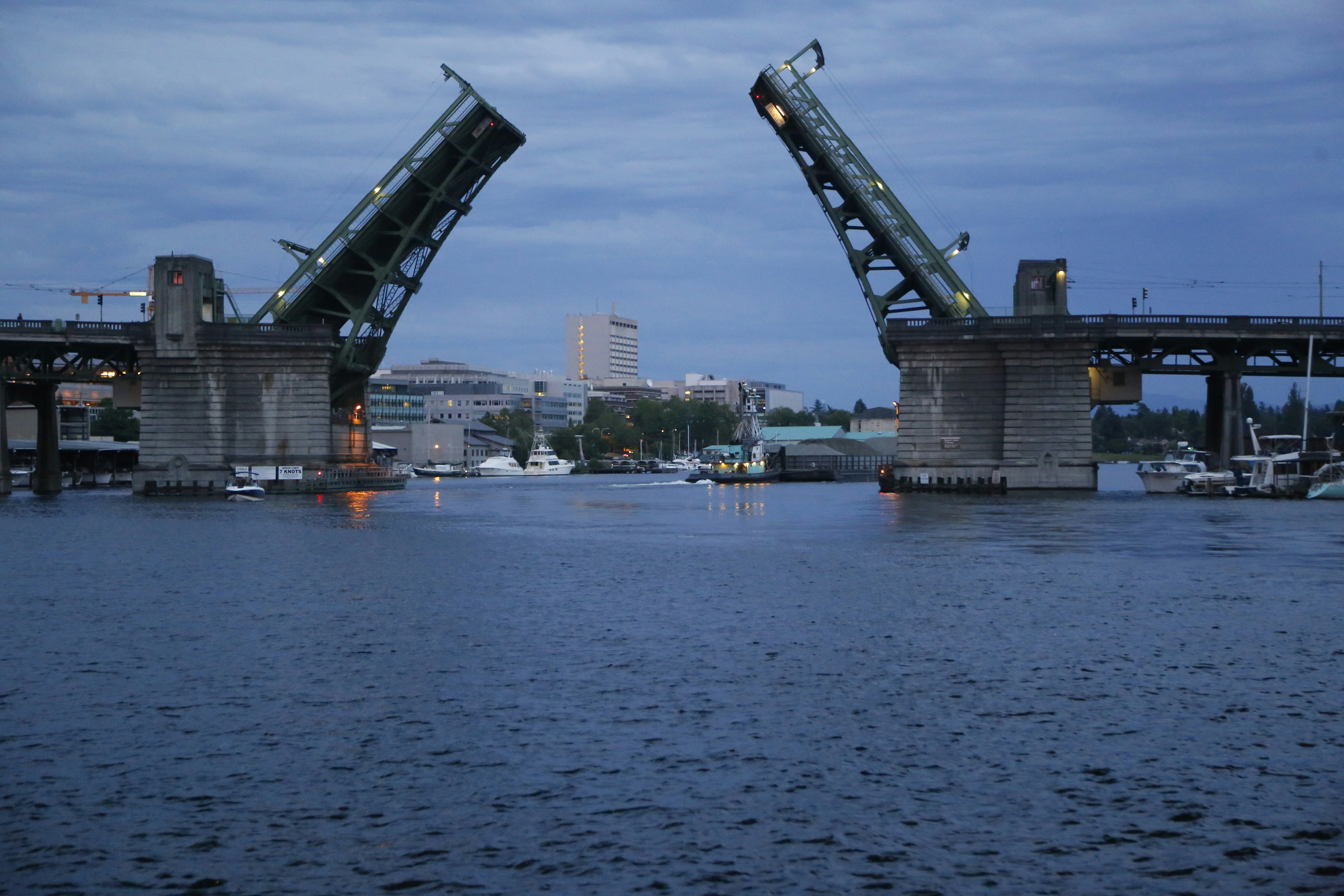 Drawbridge lifting over calm waters with city skyline in the background during twilight.