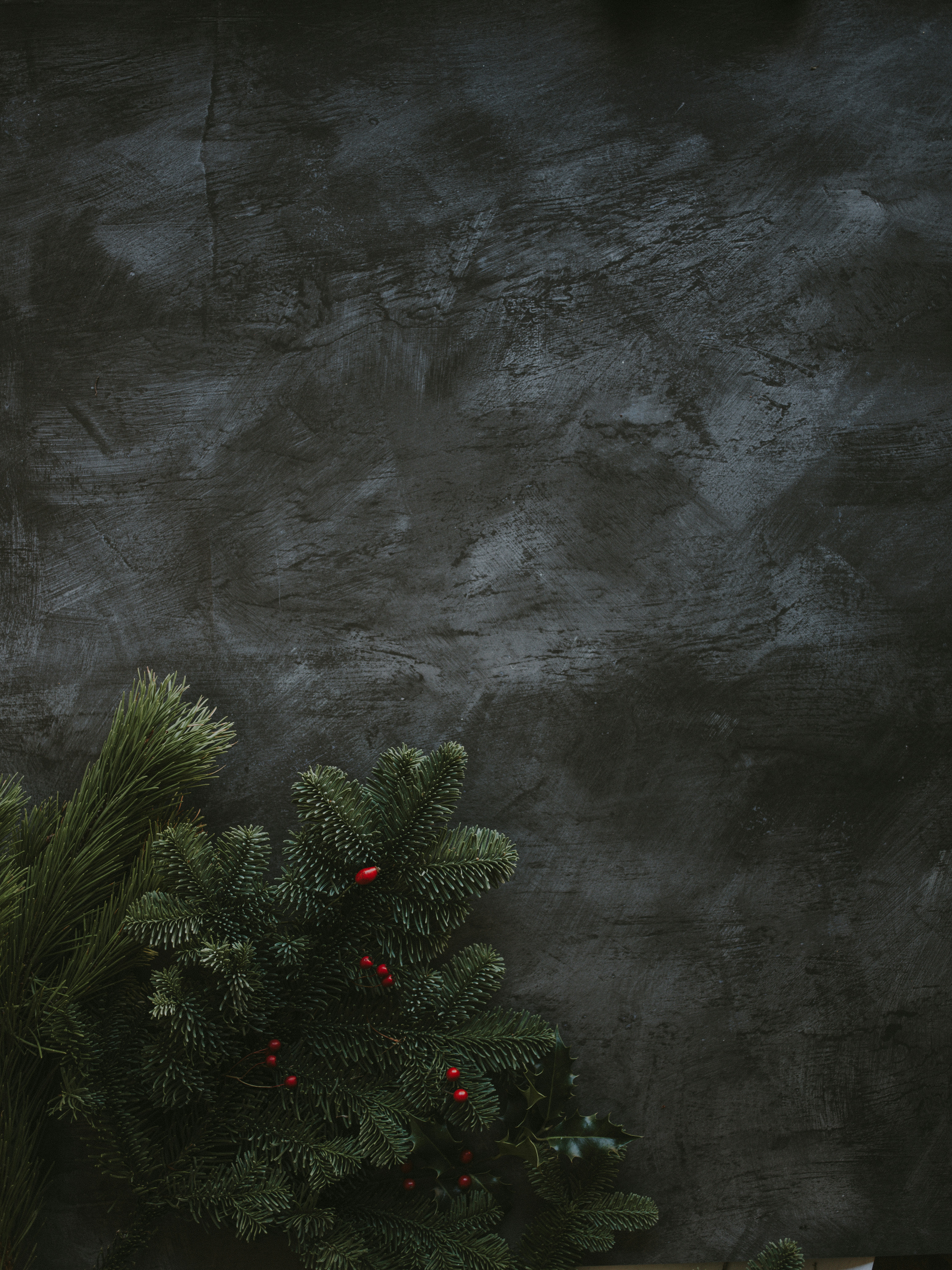 A visually striking flatlay featuring lush evergreen branches adorned with vibrant red berries against a textured dark gray background. The rich green hues of the foliage contrast beautifully with the subtle, muted tones of the backdrop, creating a serene and festive atmosphere. The composition highlights the natural beauty and simplicity of the holiday season.