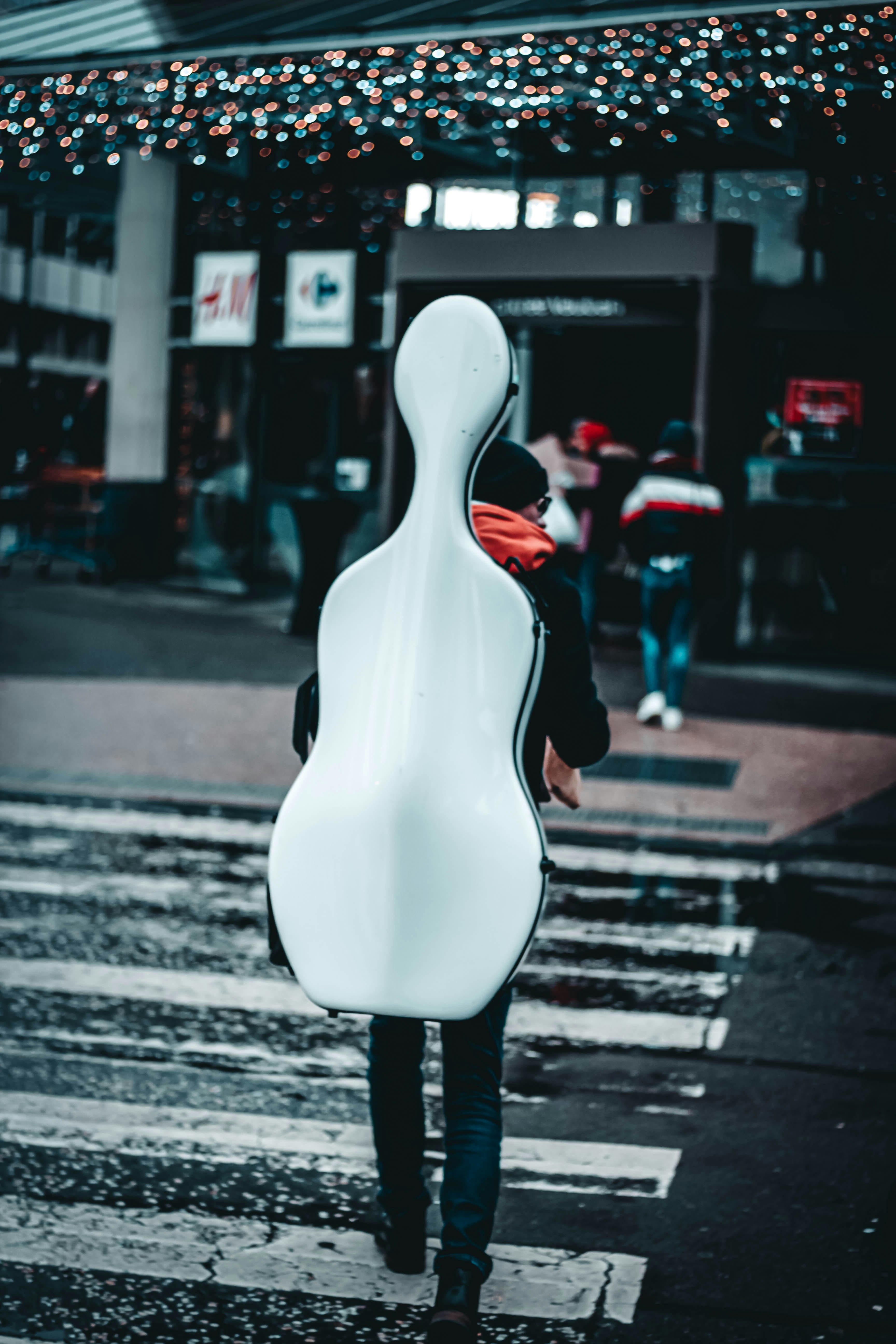 A musician traverses a busy street, carrying a white cello case on their back. The scene captures the essence of urban life intertwined with the passion for music.