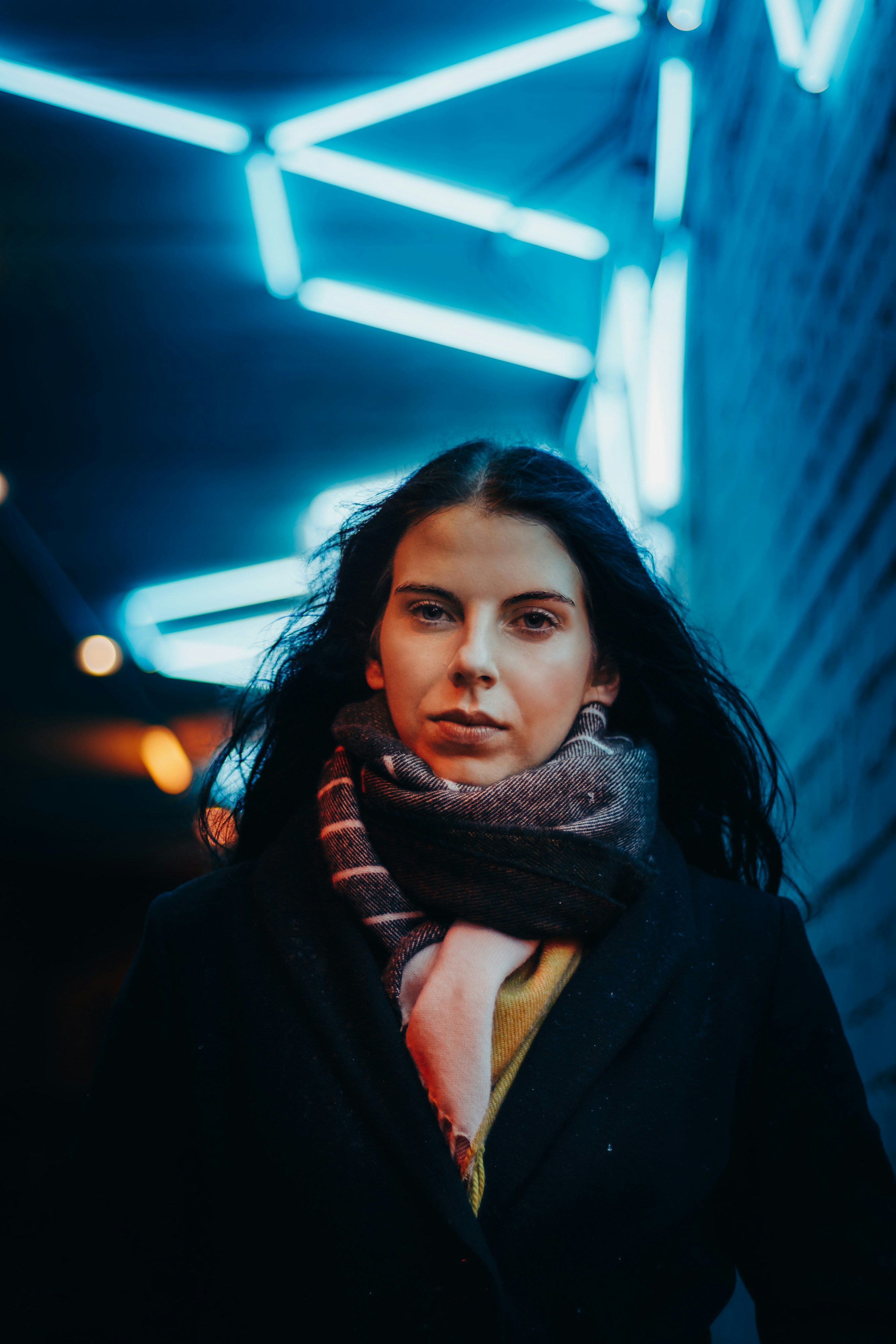 A young woman stands confidently against a backdrop of glowing neon lights, wrapped in a cozy scarf. The scene captures a modern urban atmosphere.