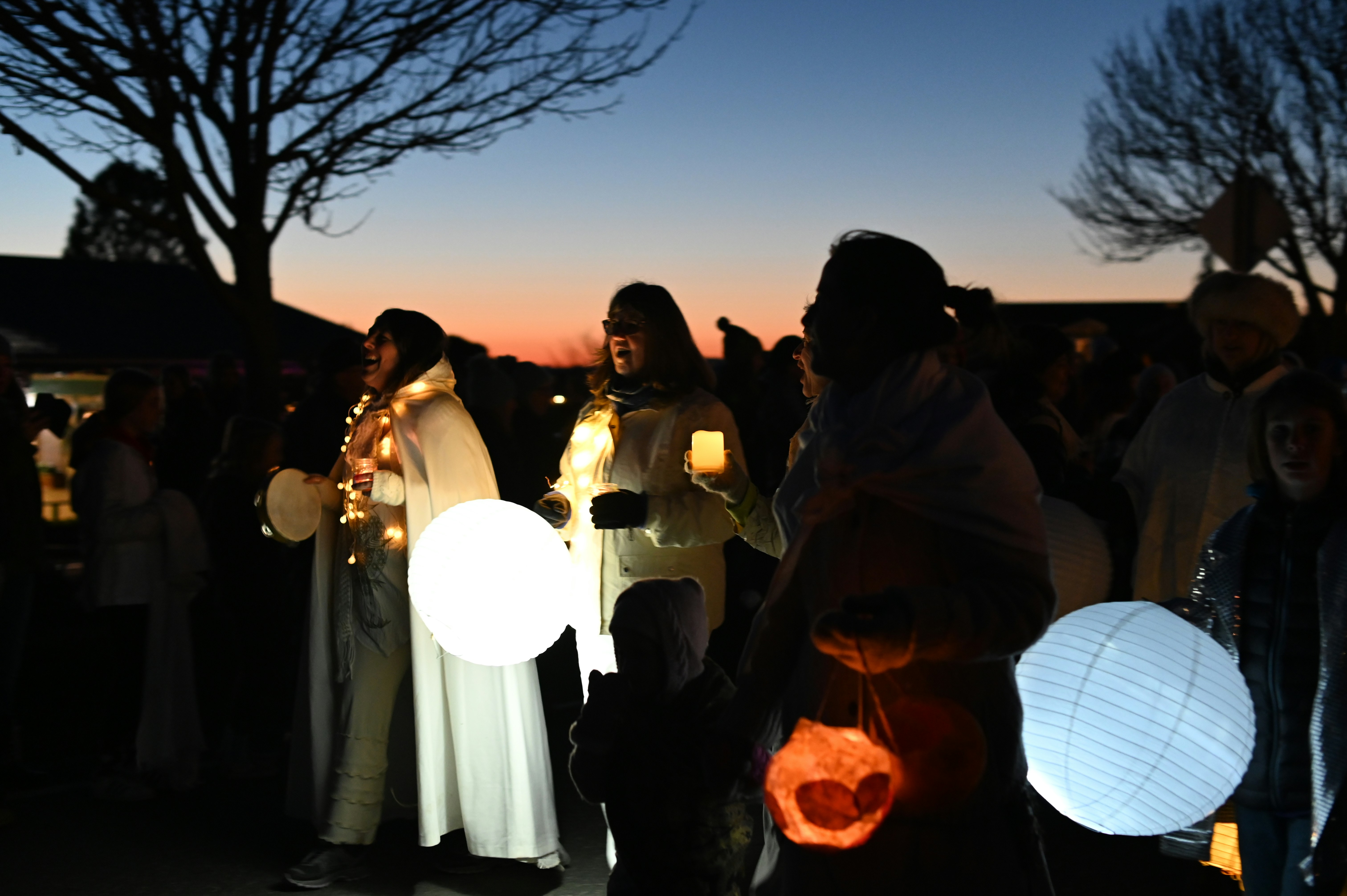 Participants in cloaks carry glowing lanterns during an evening parade against a twilight sky.