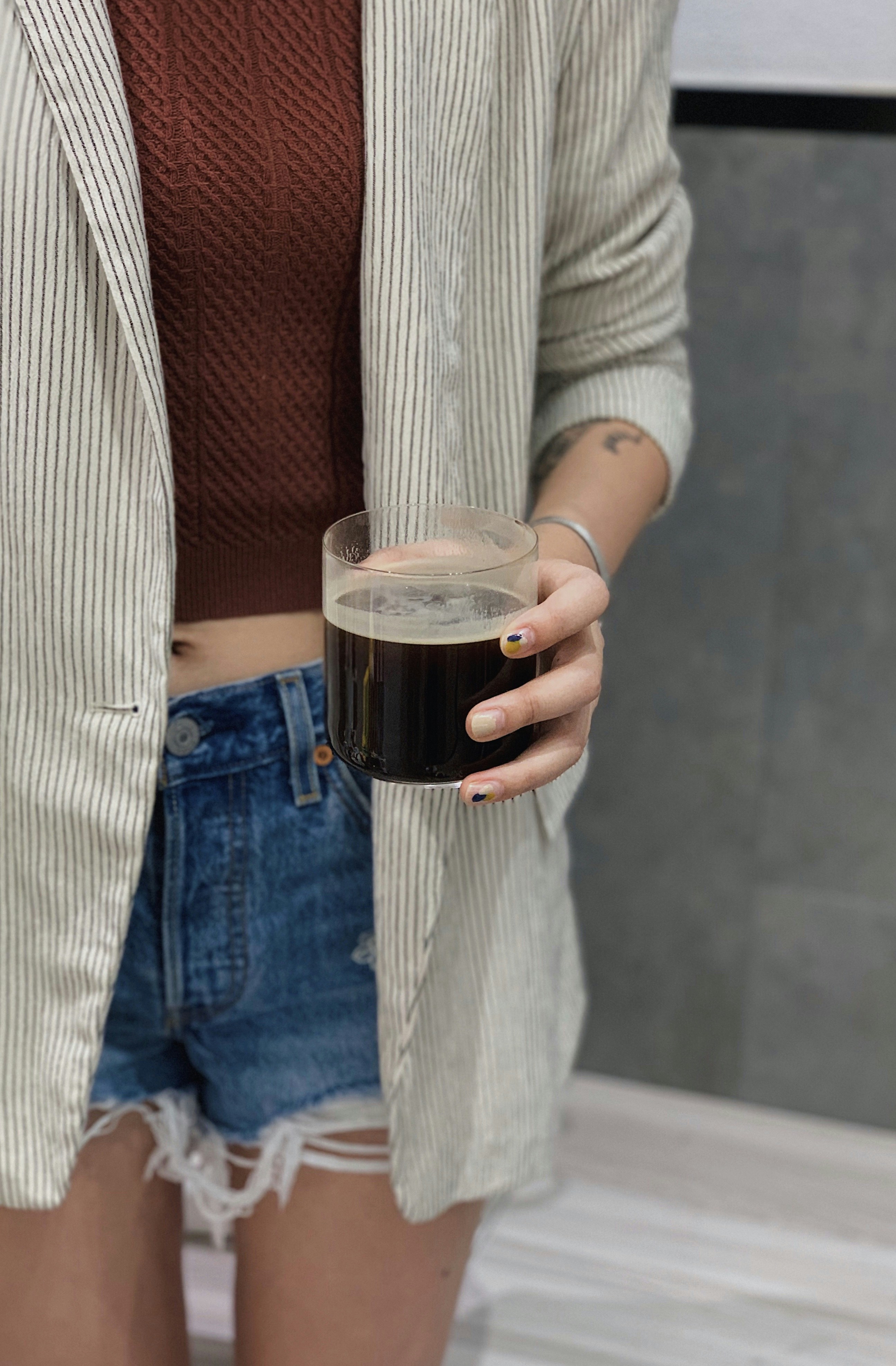 Individual holding a glass of dark beverage while dressed in a casual outfit featuring a striped blazer and denim shorts.