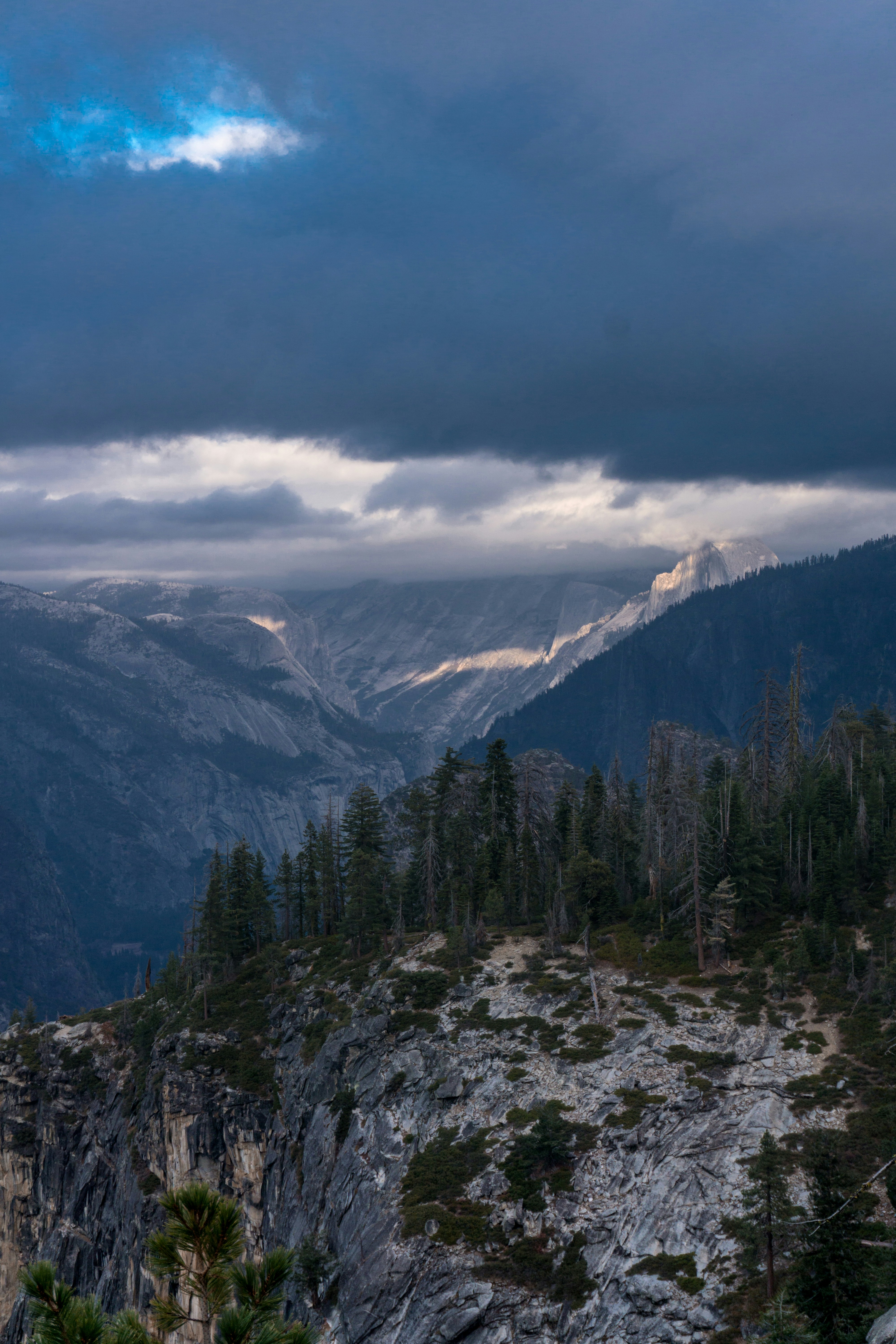 Dramatic mountain landscape with dense forests and rugged cliffs under a moody sky, revealing distant peaks. The interplay of light and shadow adds depth to the scene.