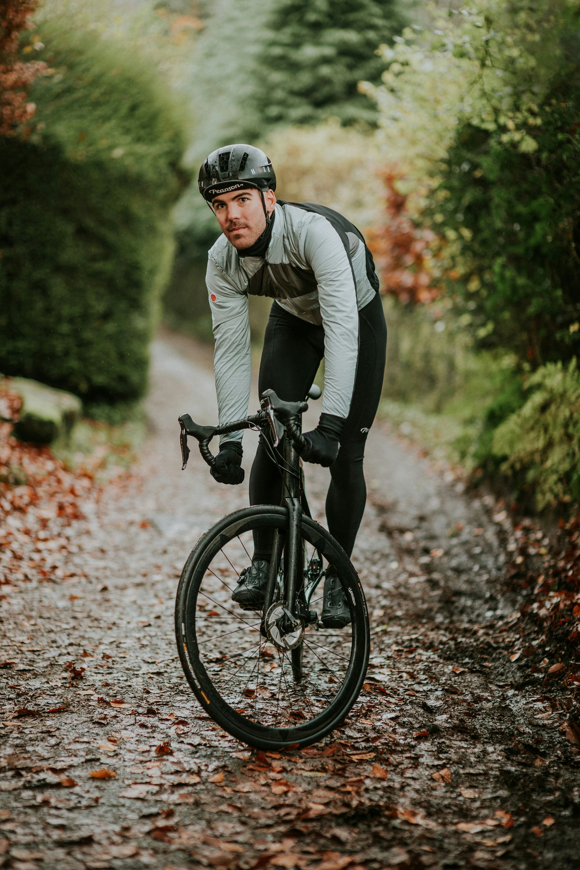 Cyclist in protective gear rides along a leaf-strewn path surrounded by dense greenery.