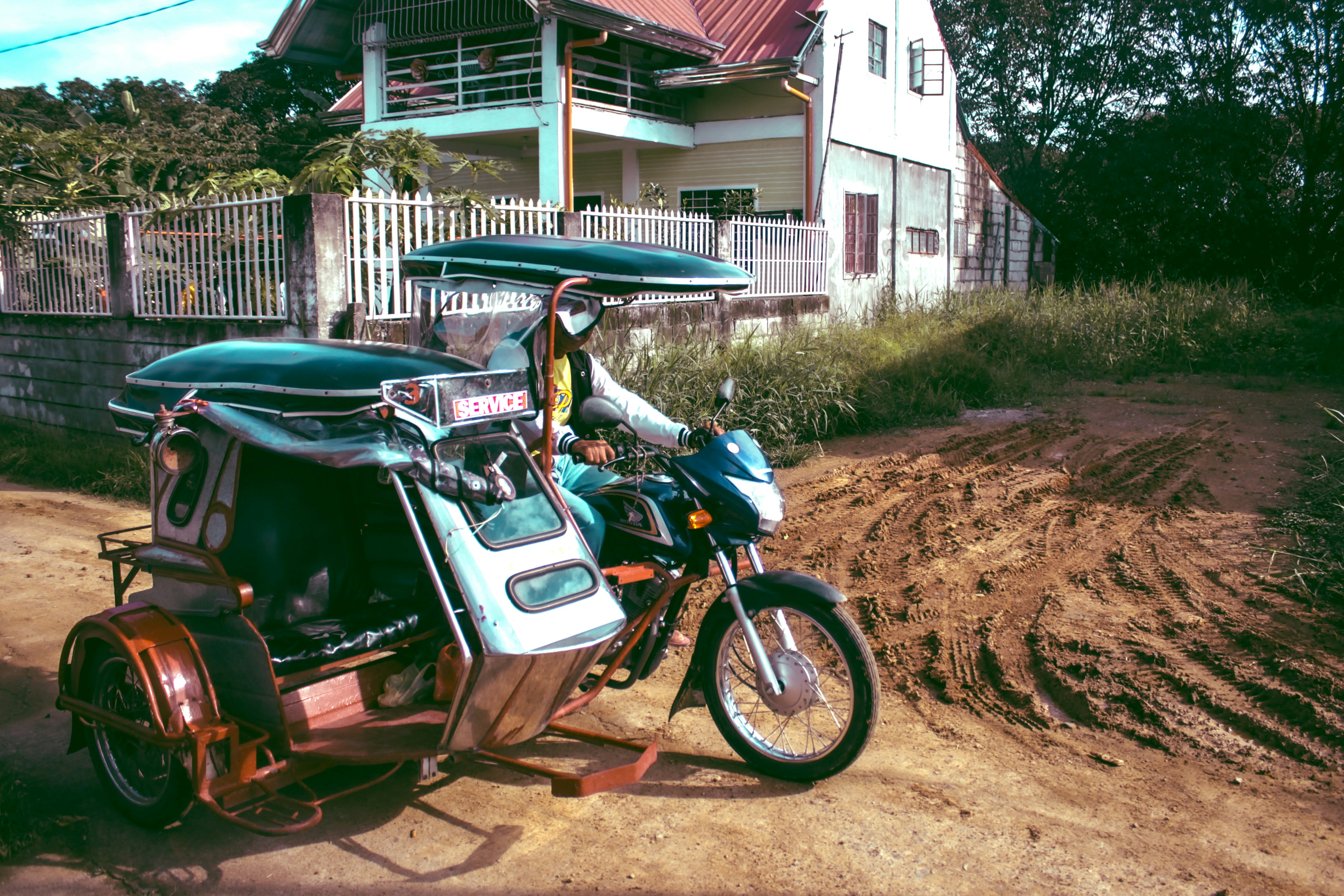 Gray tricycle parked on a dirt road beside a house with a wraparound balcony.