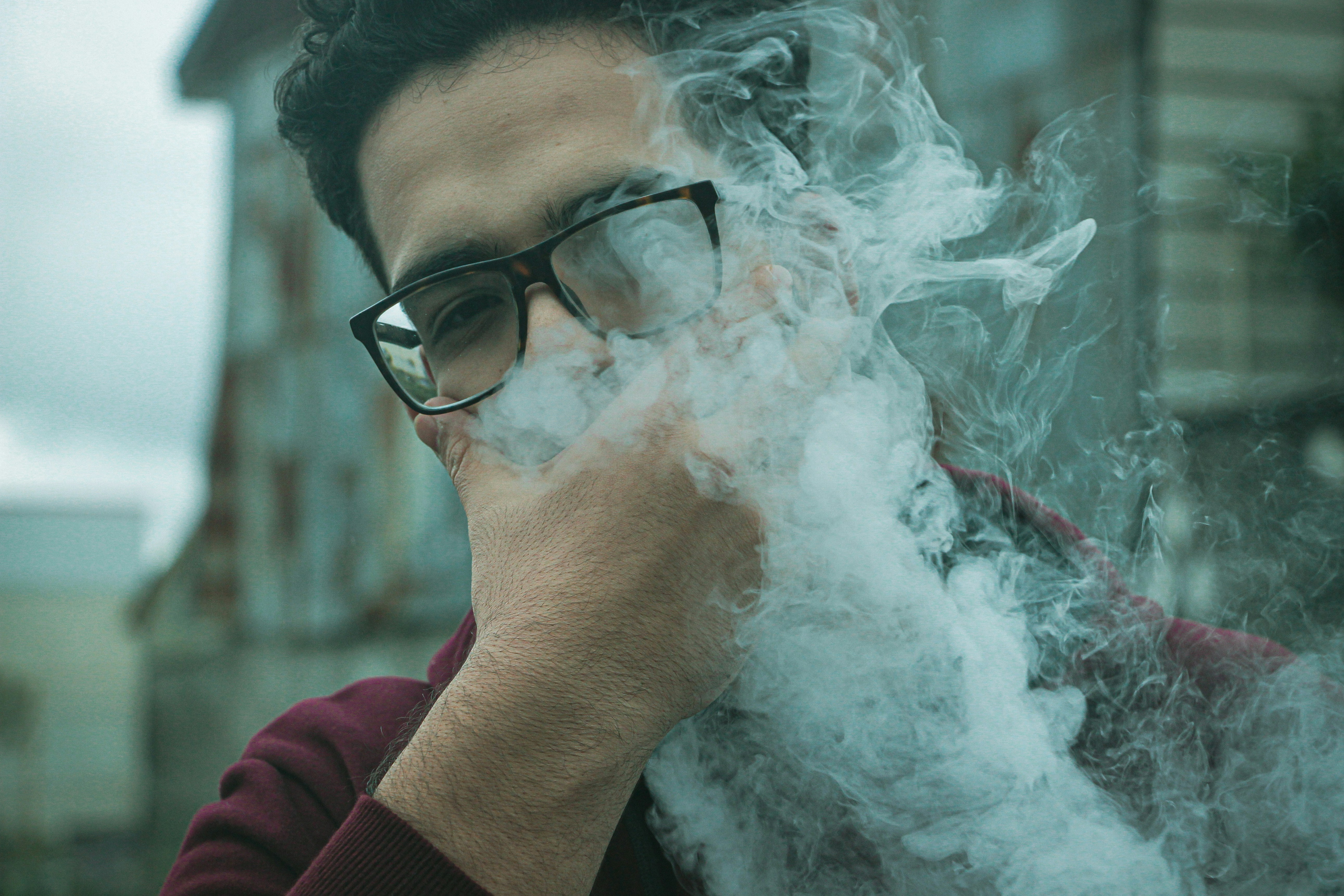 Person with glasses enveloped in a cloud of smoke outdoors.
