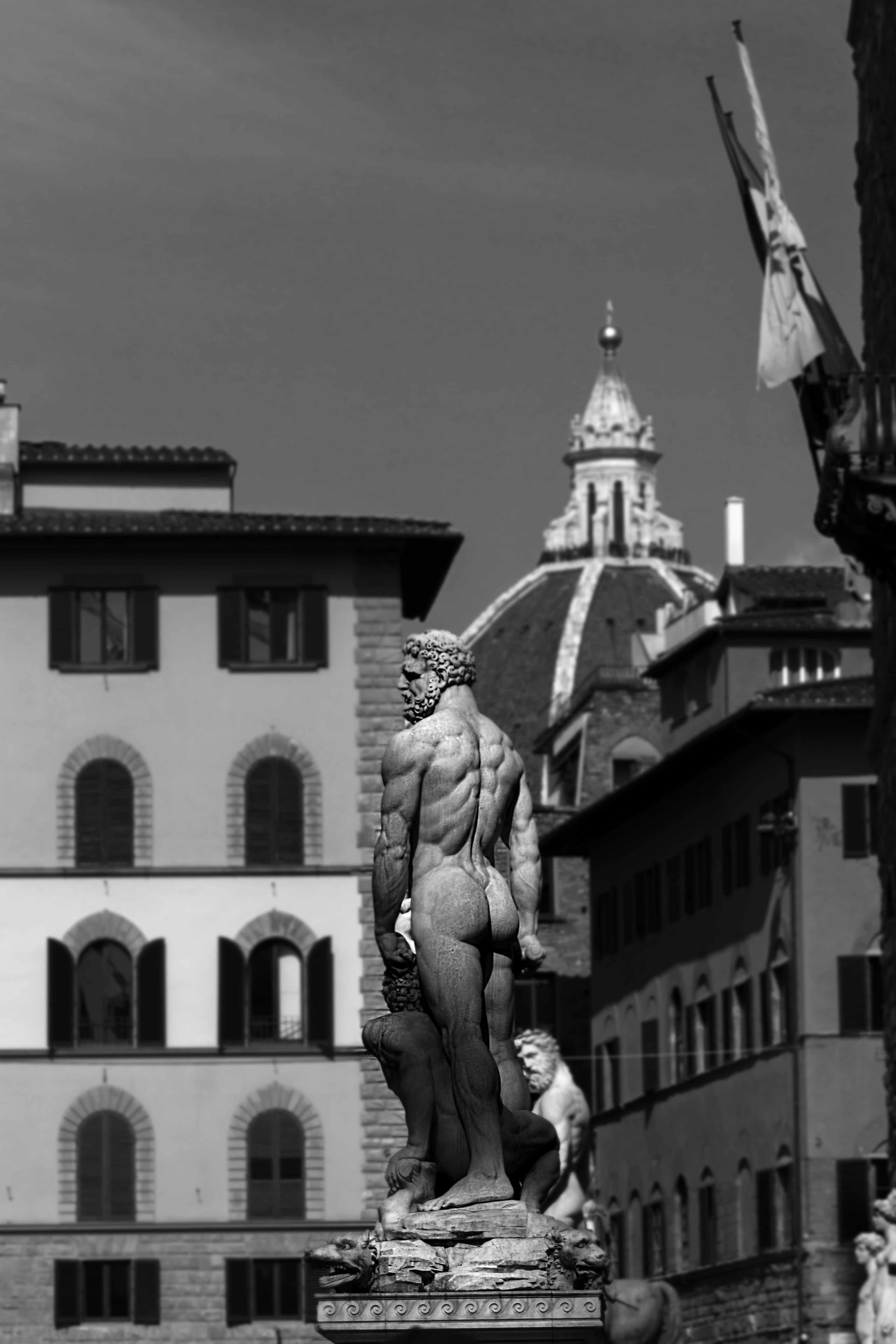 Grayscale photo of a muscular statue in an Italian piazza with historic buildings and a domed structure in the background.